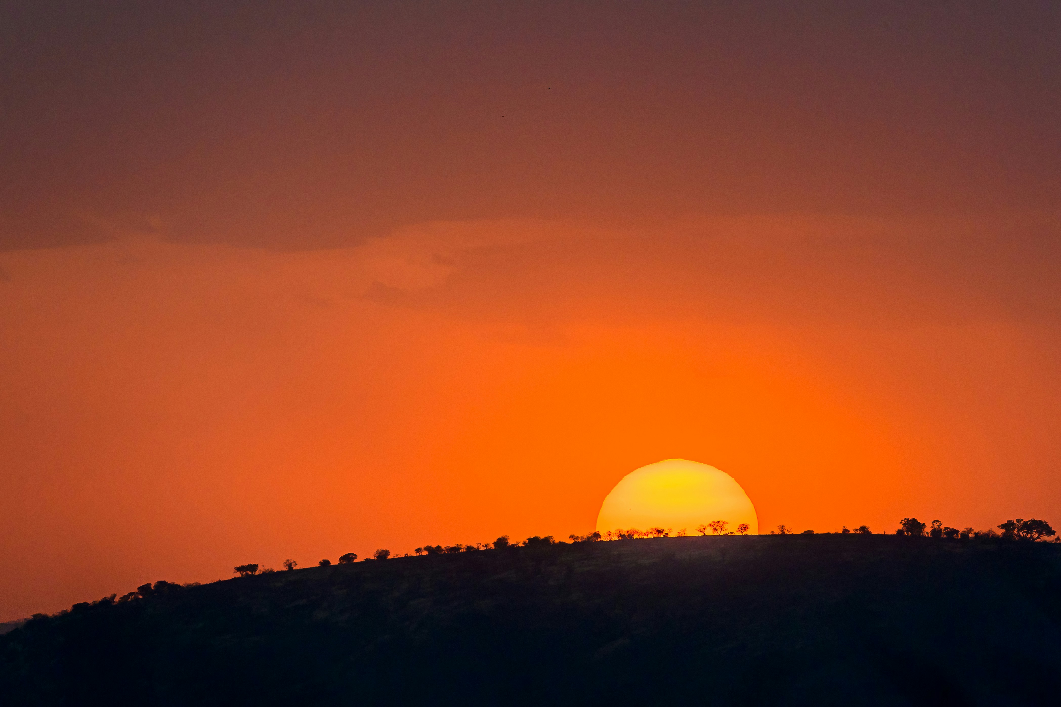 Warm sunset over a dark hill silhouette with the sun half-embedded on the horizon; a minimal landscape emphasizing the gradient sky and soft silhouettes.