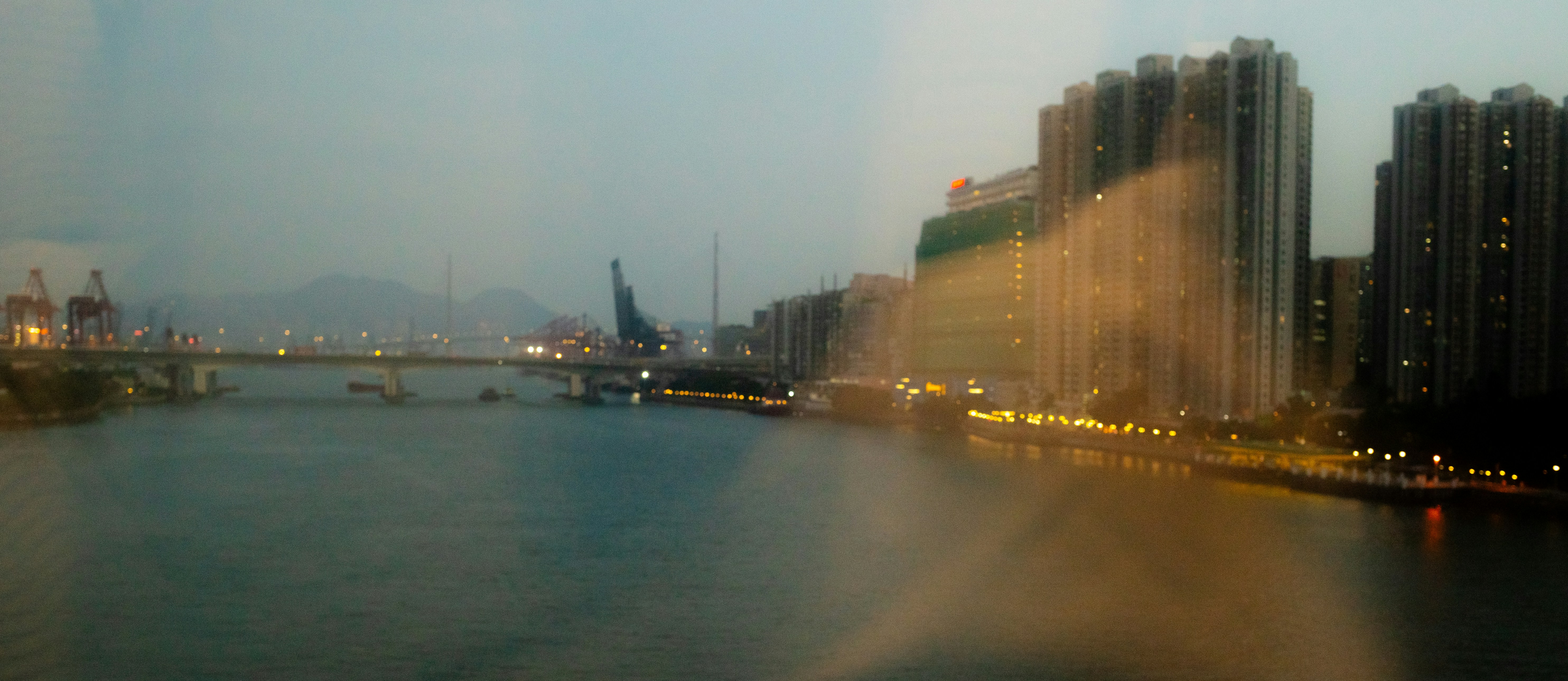 A calm evening view of Hong Kong’s waterfront, with high-rise residential towers, glowing city lights, and reflections on the water. The shoreline promenade and urban skyline create a quiet but dense city atmosphere.