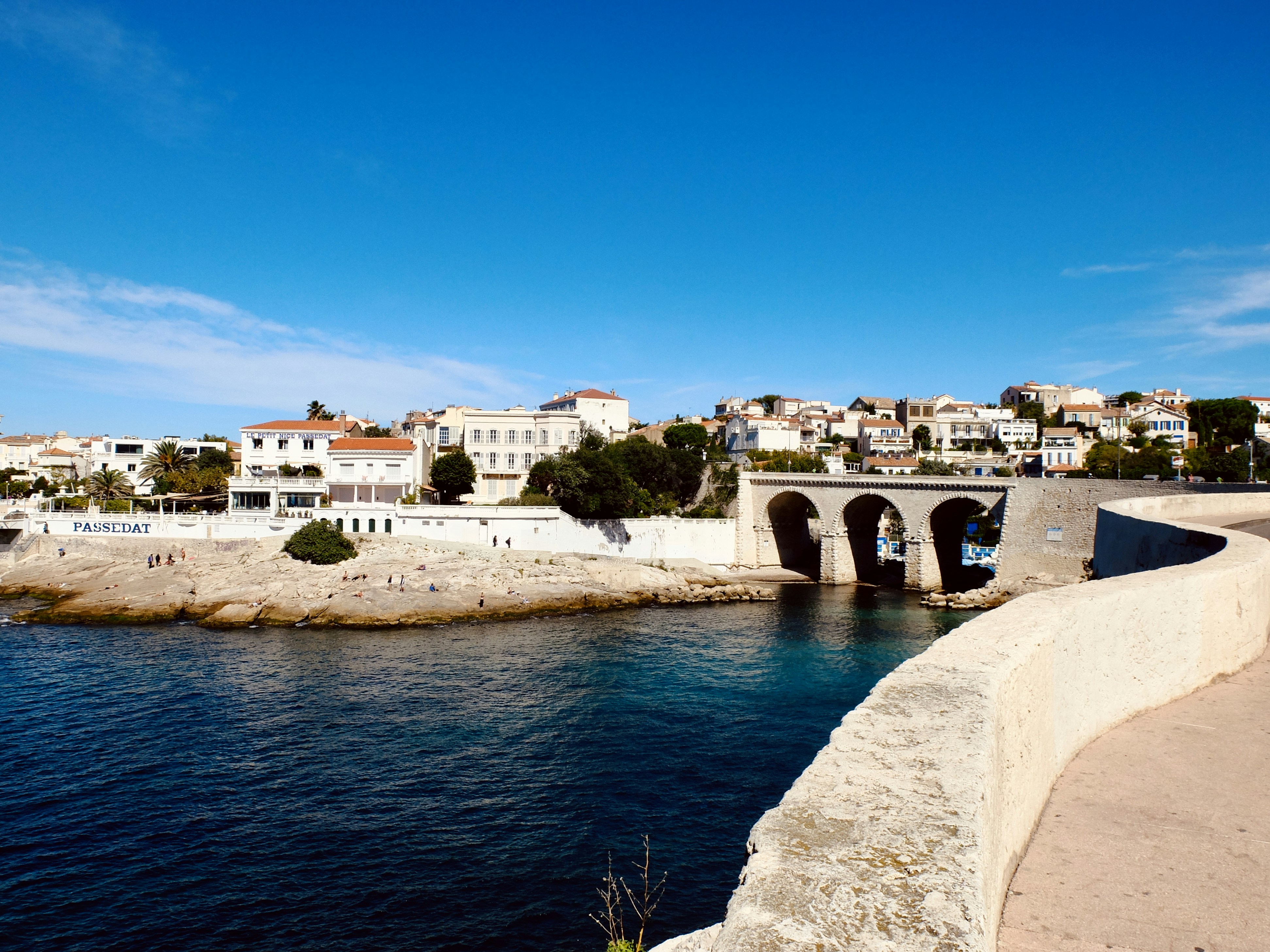 Sunny coastal town vista with a stone bridge arching over calm blue water and a curved seawall in the foreground.