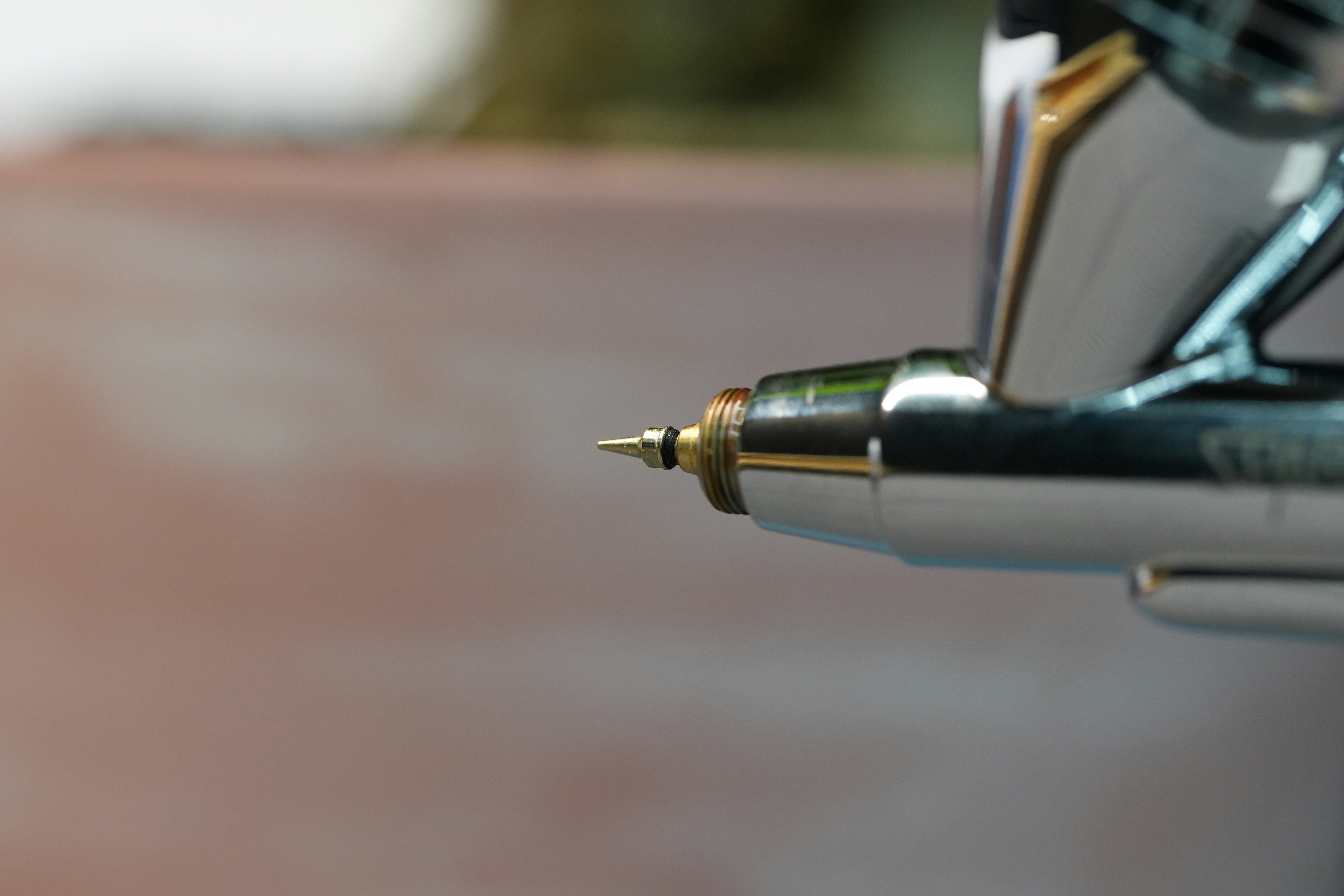 Close-up of a pen nib and tip extended from a metallic pen body; blurred background suggesting a workbench or desk, with emphasis on the tool tip and chrome detailing.