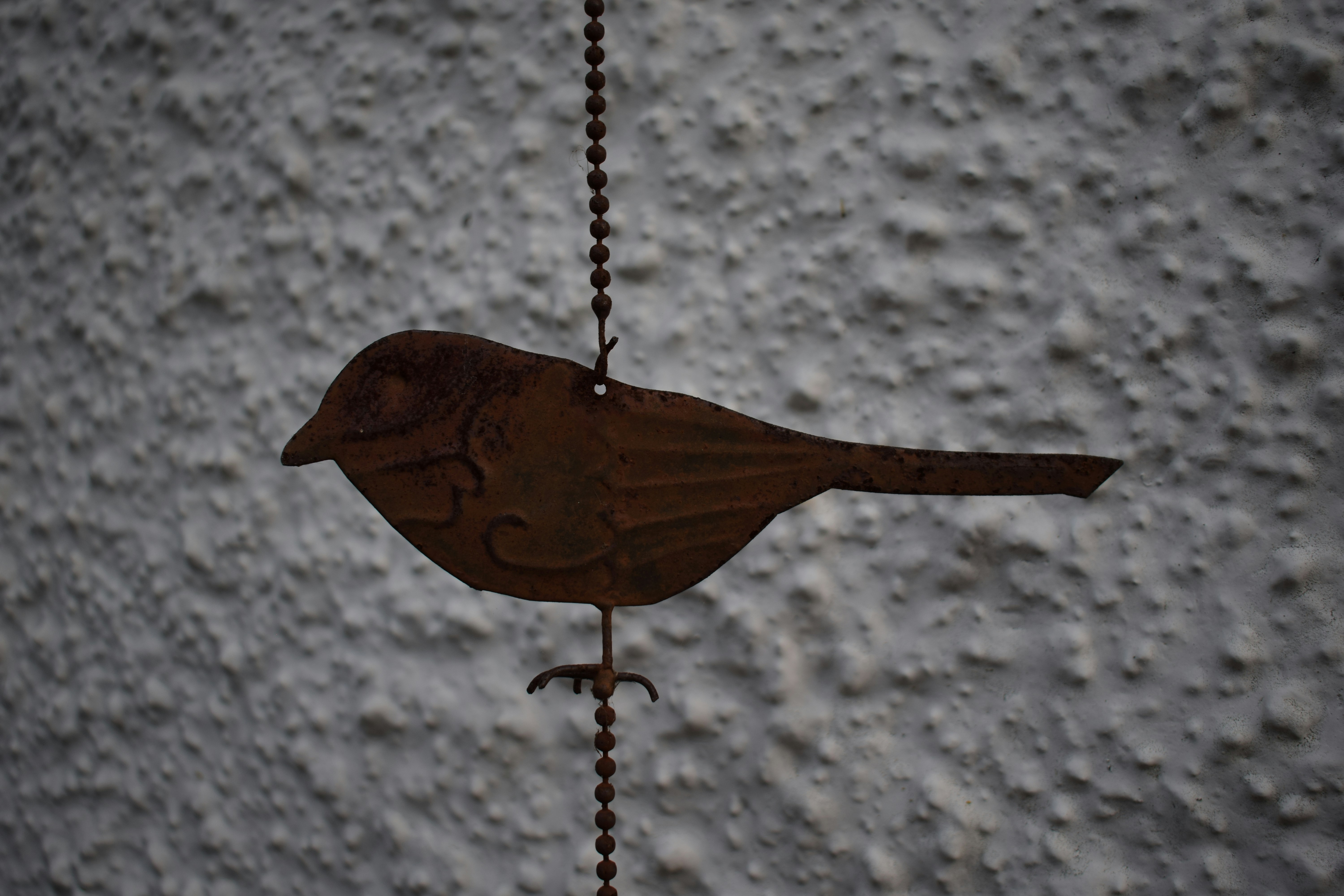 Rusty bird ornament suspended on a beaded chain against a textured wall