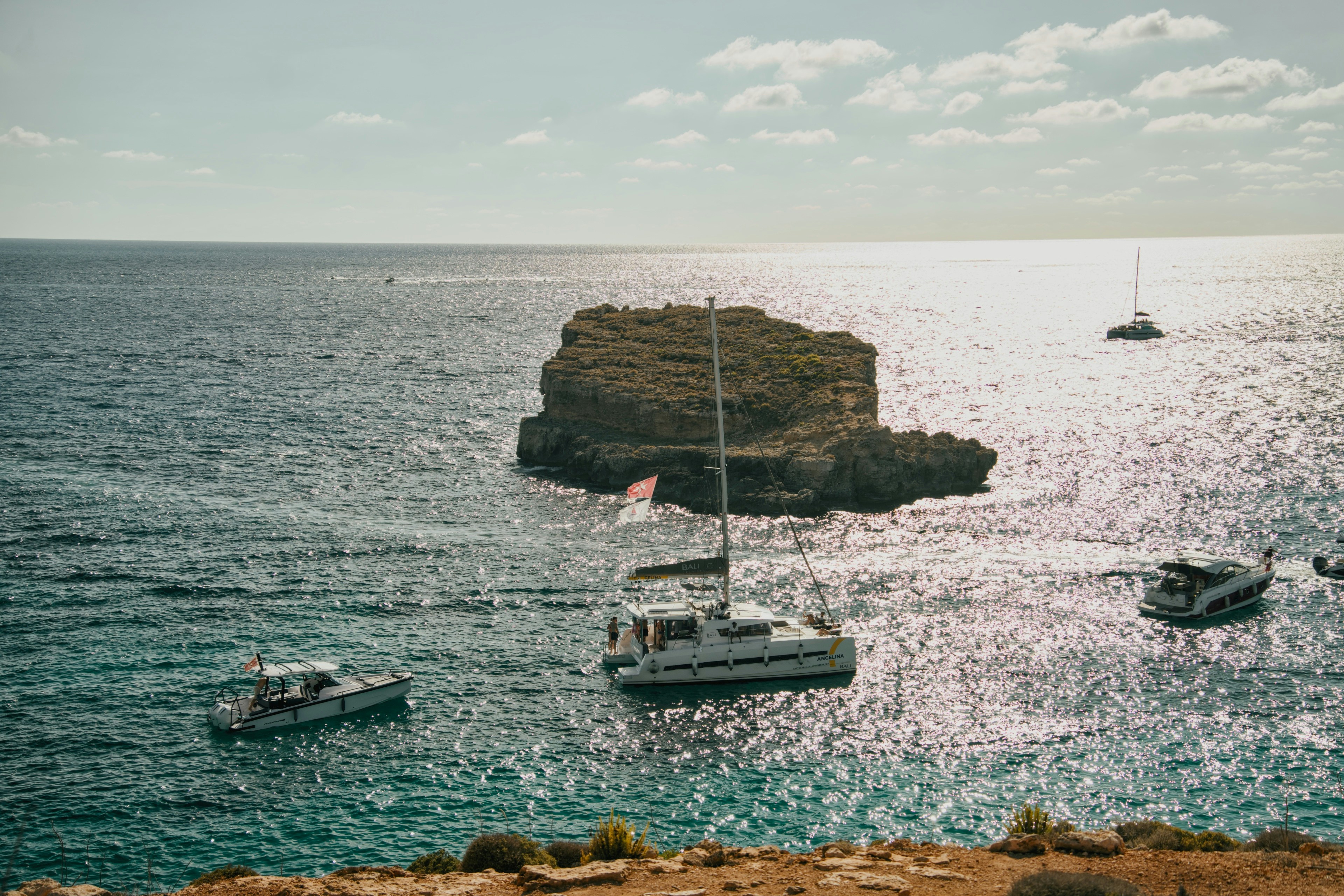 Small fleet of boats clusters around a rocky outcrop bathed in late-afternoon light, with sparkling water and a distant sailboat; a coastal scene emphasizing maritime activity and tranquil seascape.