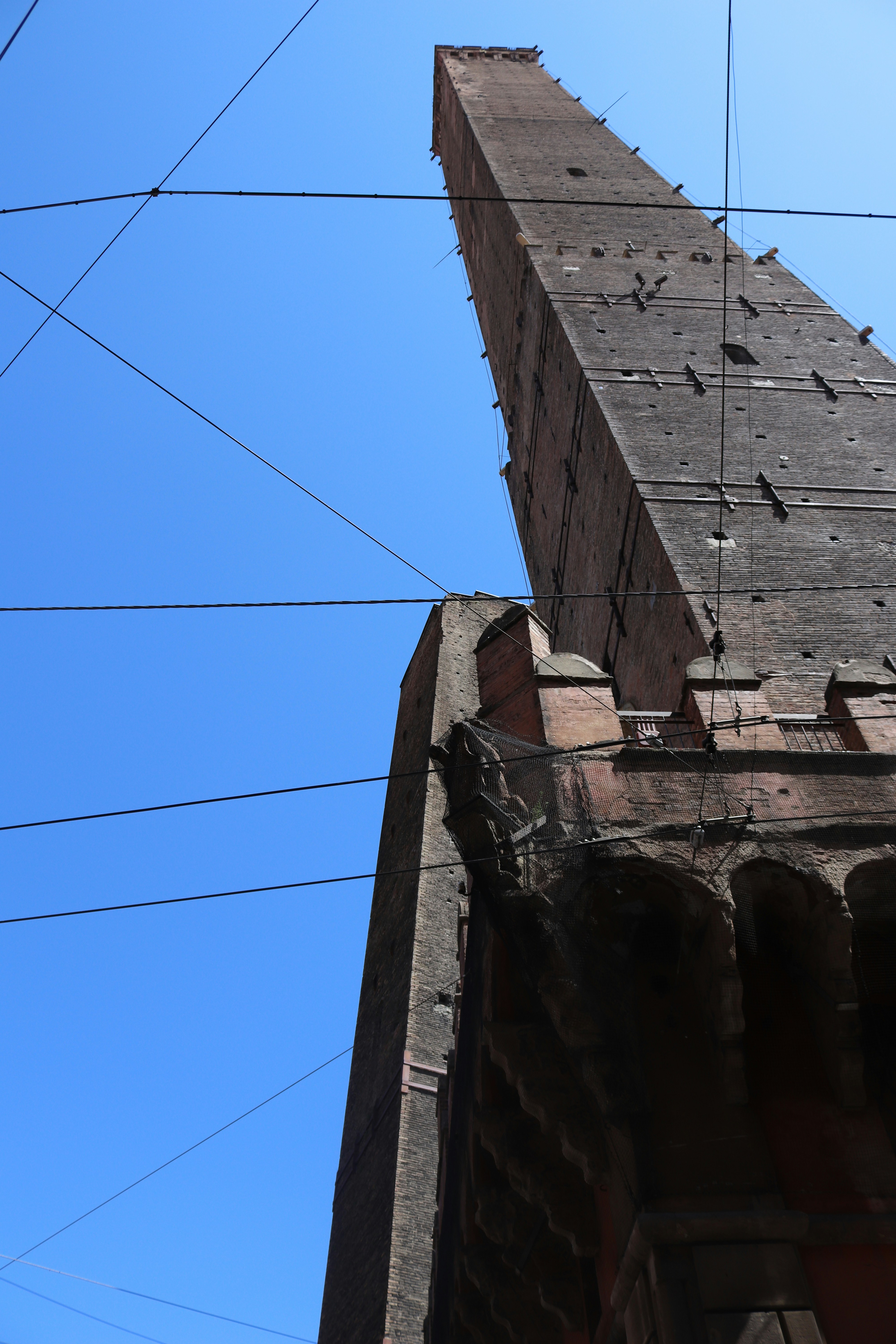 Tall brick tower rising into a blue sky with intersecting power lines across the frame; architectural detailing visible at the base adds texture to the composition.