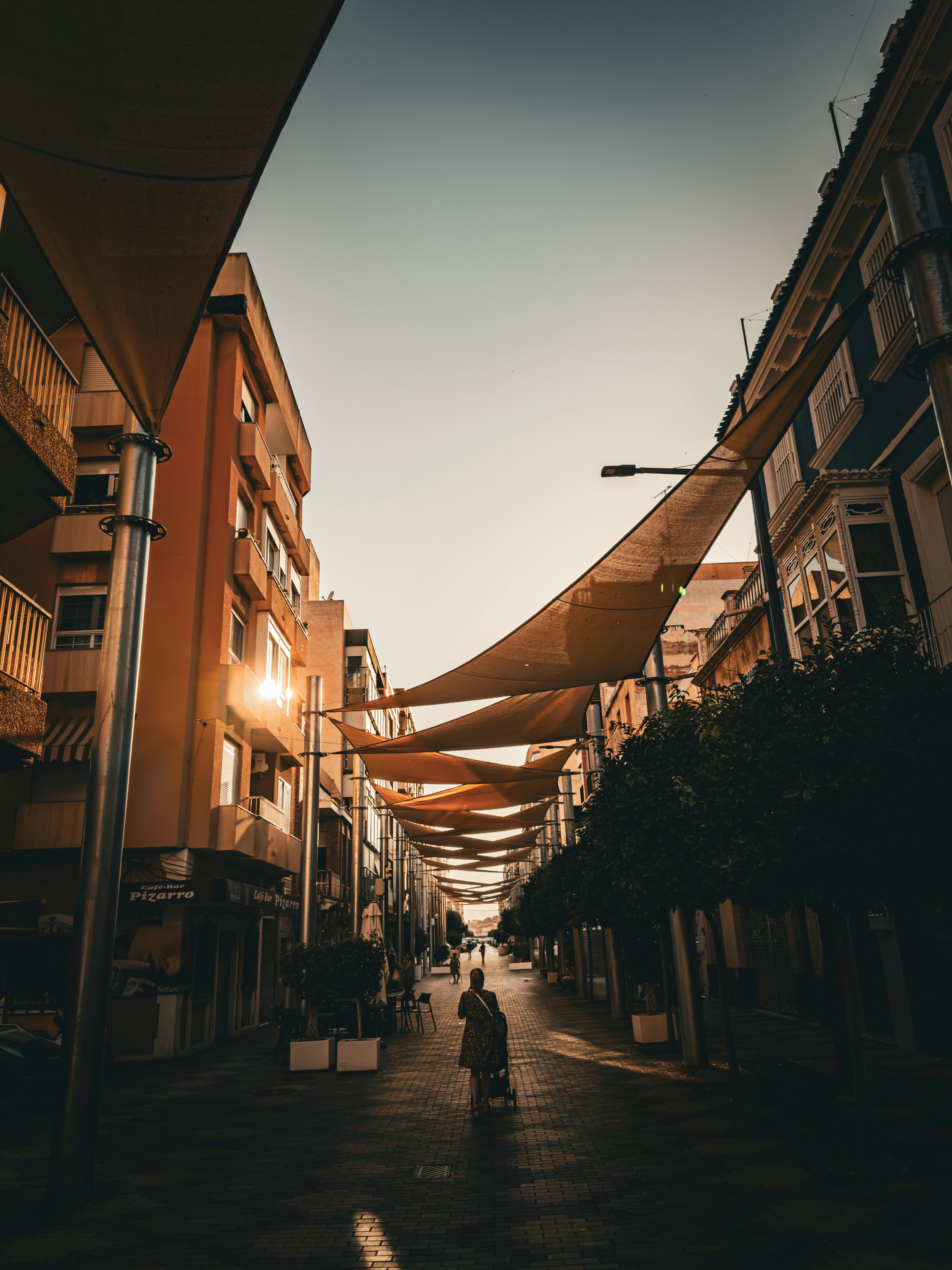 A sunlit urban pedestrian street with fabric canopies stretched above, casting warm tones on buildings and a lone figure pushing a stroller down the center. A logo or storefronts peek along the sides, emphasizing depth and mood.