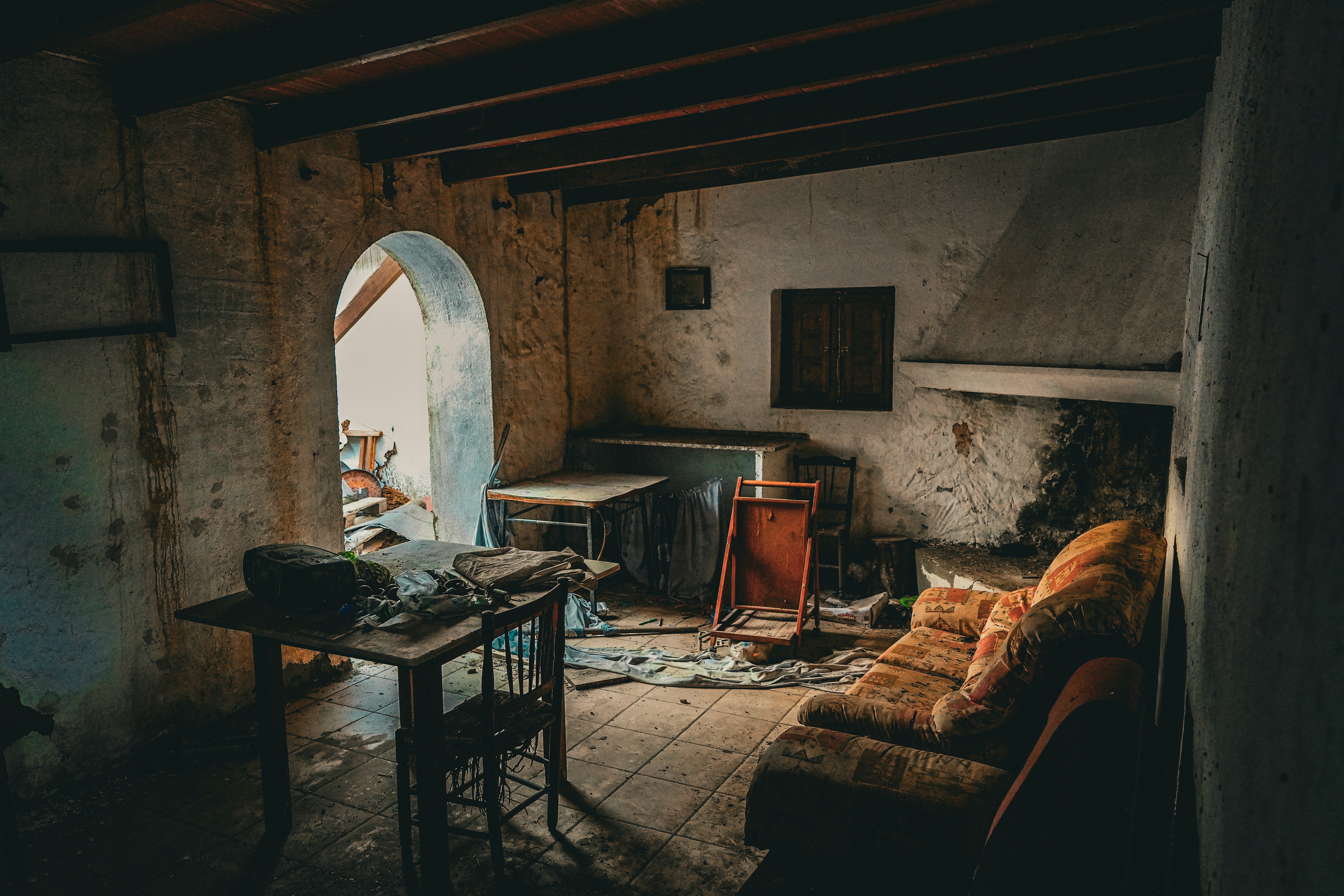 Dusty, dim living space with an arched doorway letting in muted light; worn sofa, scattered furniture, and weathered walls convey a deserted, vintage interior. (photograph)