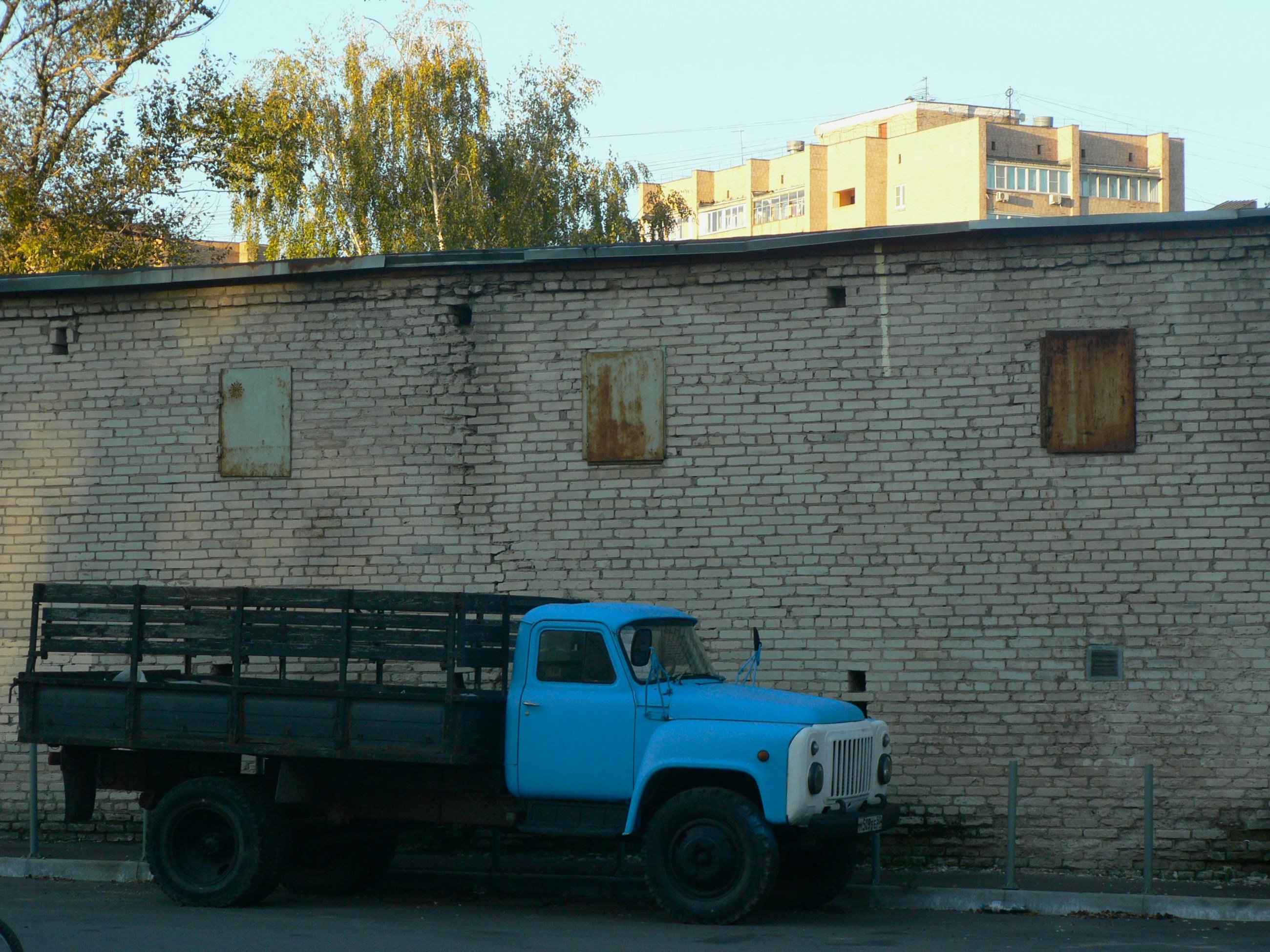 A classic Soviet-era flatbed truck, painted in a bright blue with a white grille, is parked alongside a long, weathered white brick wall. The truck features a wooden slatted cargo bed, black tires, and a simple, utilitarian cabin. The brick wall behind it is marked by three rusty metal panels that appear to cover old windows or openings. In the background, a yellow multi-story building rises above the wall, and tall trees with green and yellowing leaves are silhouetted against a clear, pale sky. The scene captures a quiet, everyday moment in an urban or industrial area.