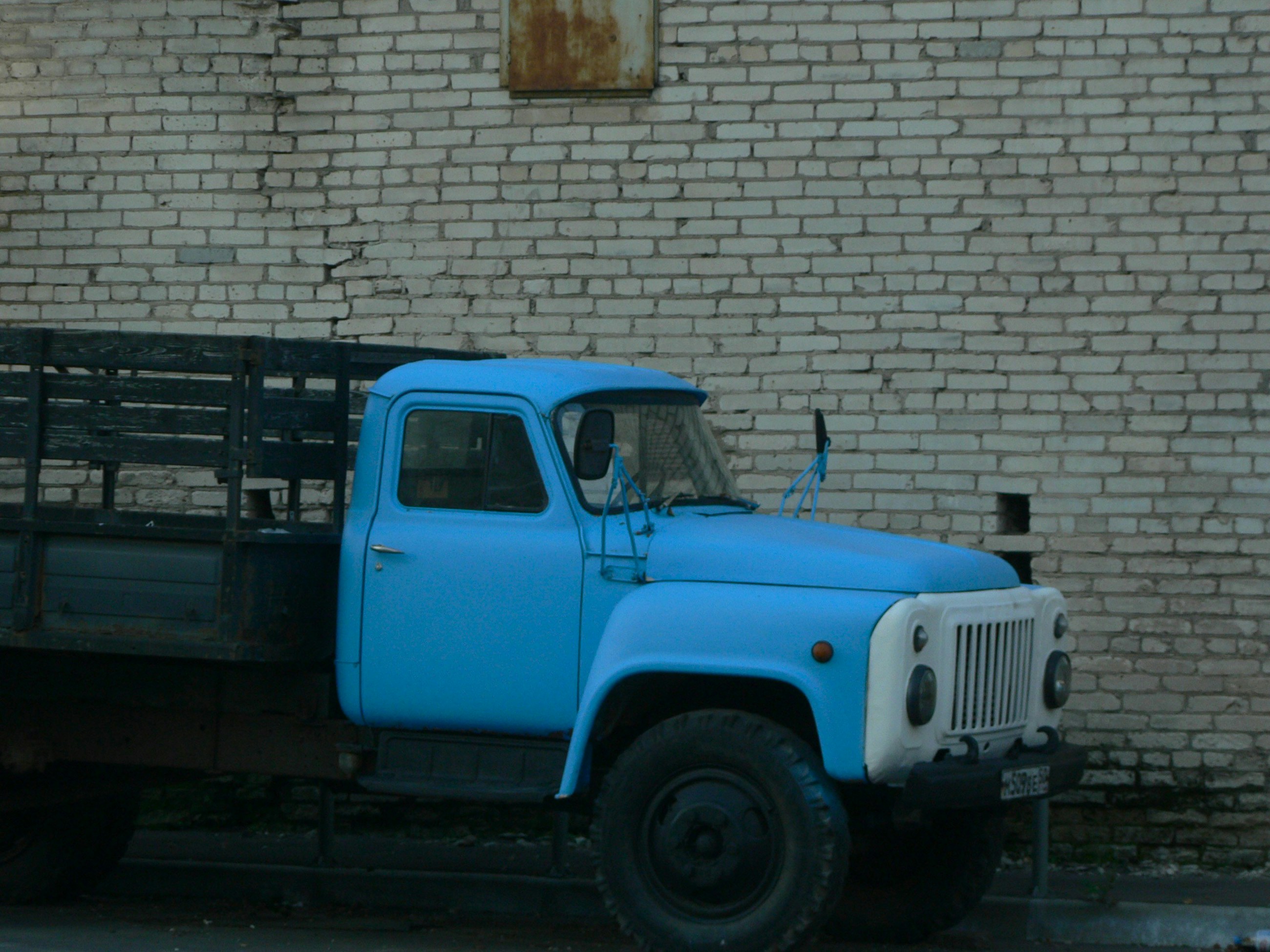 A classic Soviet-era flatbed truck, painted in a bright blue with a white grille, is parked alongside a long, weathered white brick wall. The truck features a wooden slatted cargo bed, black tires, and a simple, utilitarian cabin. The brick wall behind it is marked by three rusty metal panels that appear to cover old windows or openings. In the background, a yellow multi-story building rises above the wall, and tall trees with green and yellowing leaves are silhouetted against a clear, pale sky. The scene captures a quiet, everyday moment in an urban or industrial area.