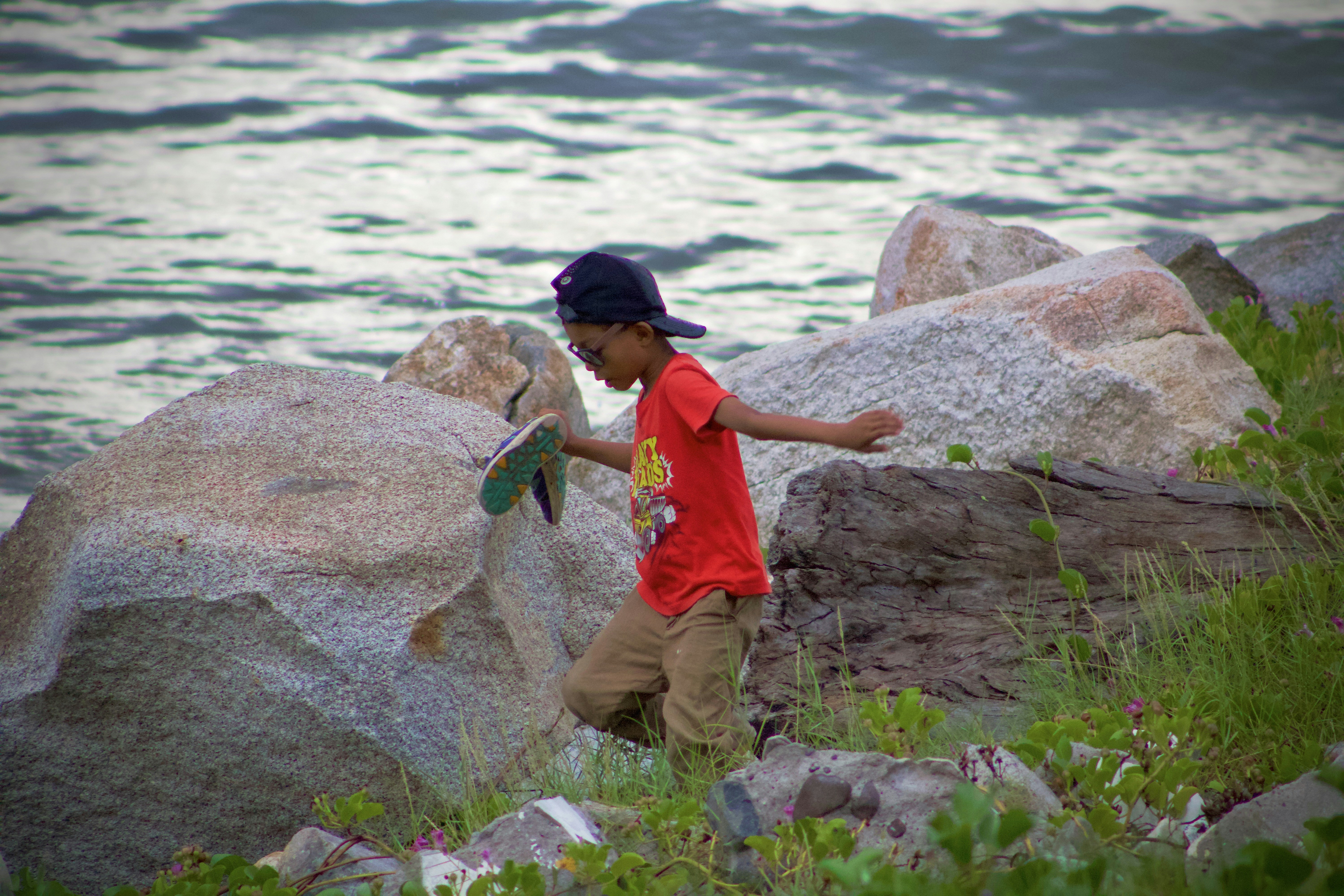 A child in a red shirt and cap navigates rocky terrain beside a calm body of water, holding a skateboard as they step between stones. The scene captures a candid moment of outdoor play and balance on uneven ground.