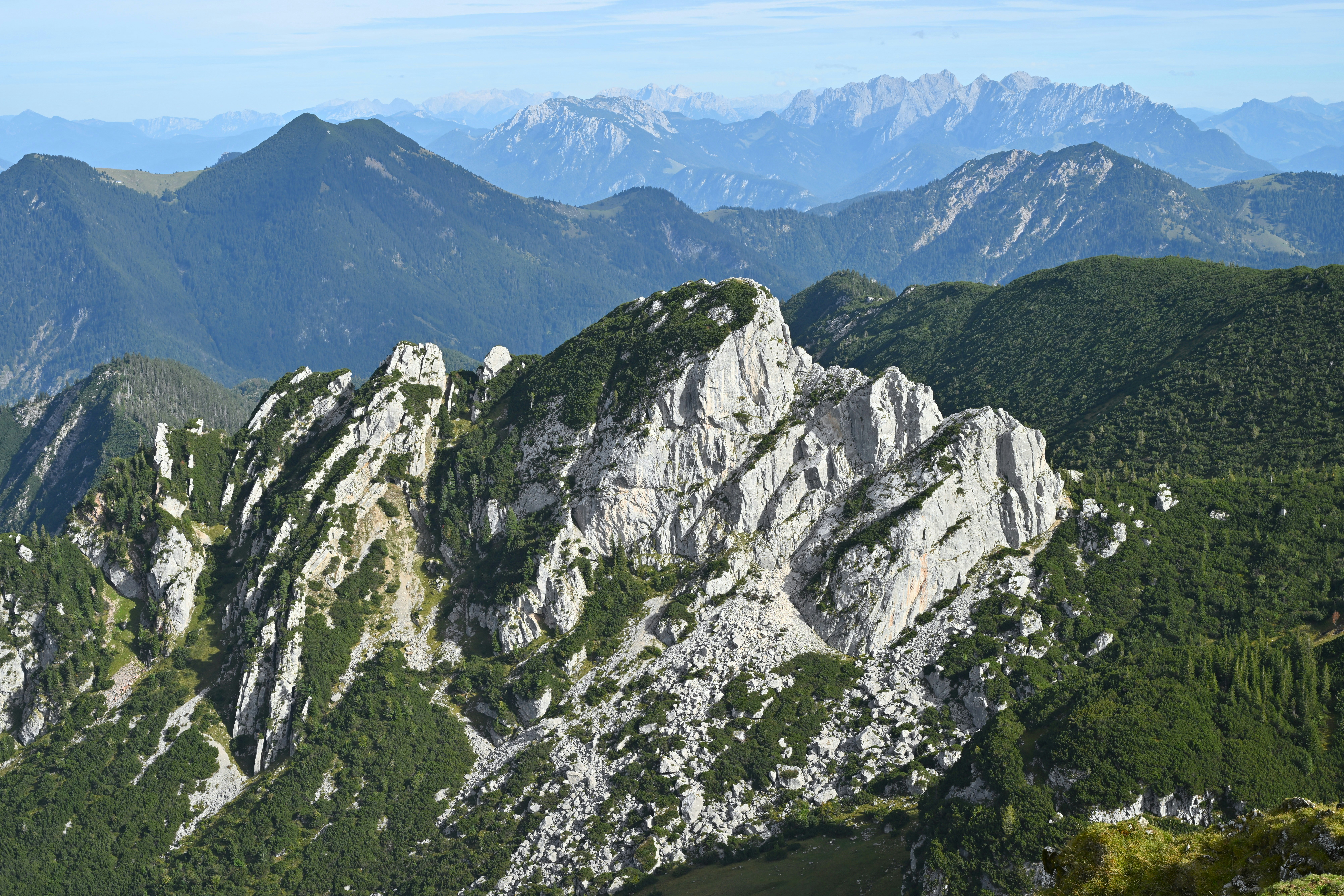 Snow-dusted jagged ridge lines cut through green mountains under a clear blue sky, with distant blue ranges creating depth.