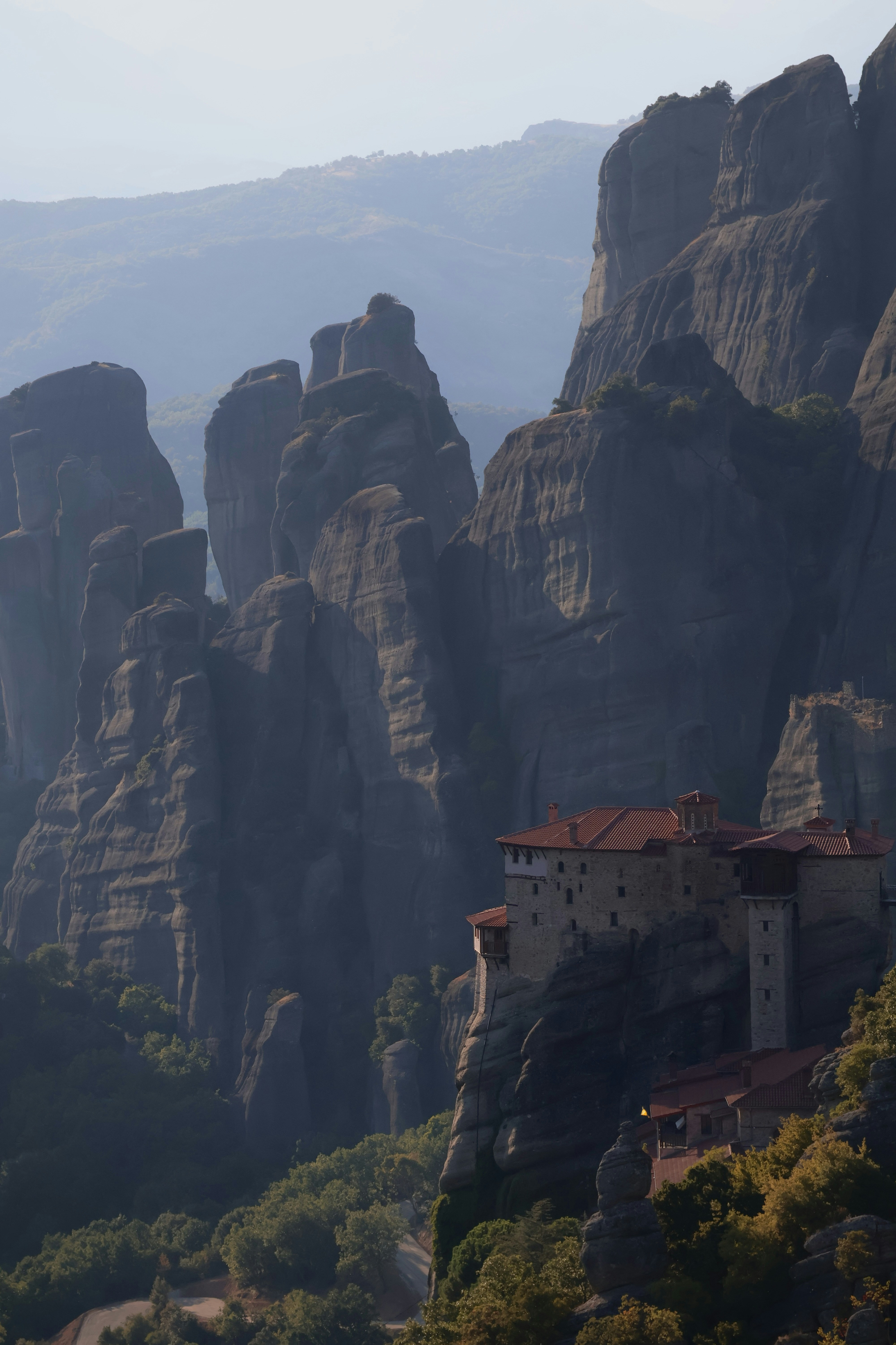 Stone fortress clings to a towering rock spire amid steep monolithic pillars, with a red-tiled roof and surrounding forest as misty mountains loom in the background.