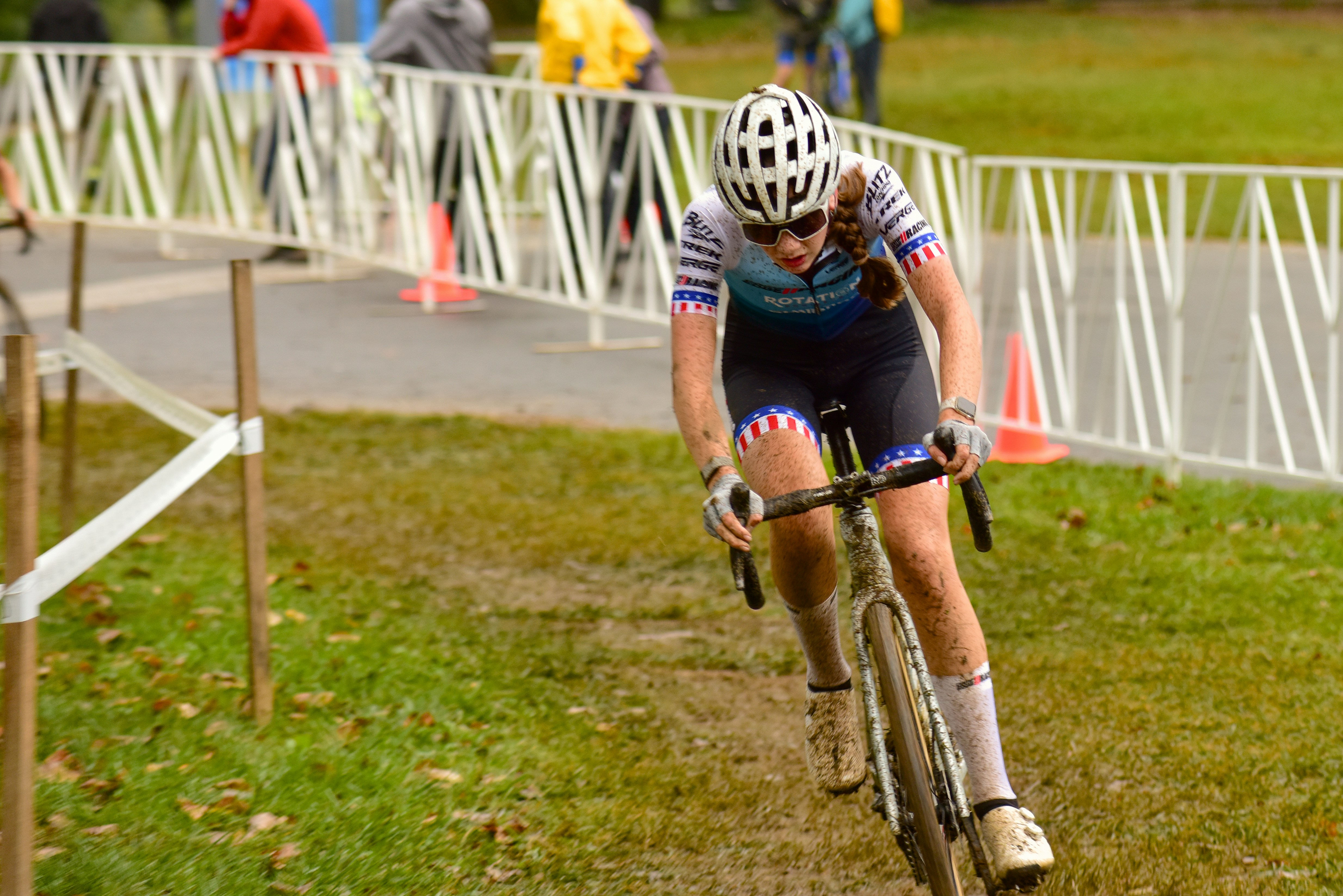 Cyclist in muddy race gear leans into a turn on a grassy track, helmeted head-down and focused, with white barriers and onlookers in the background.