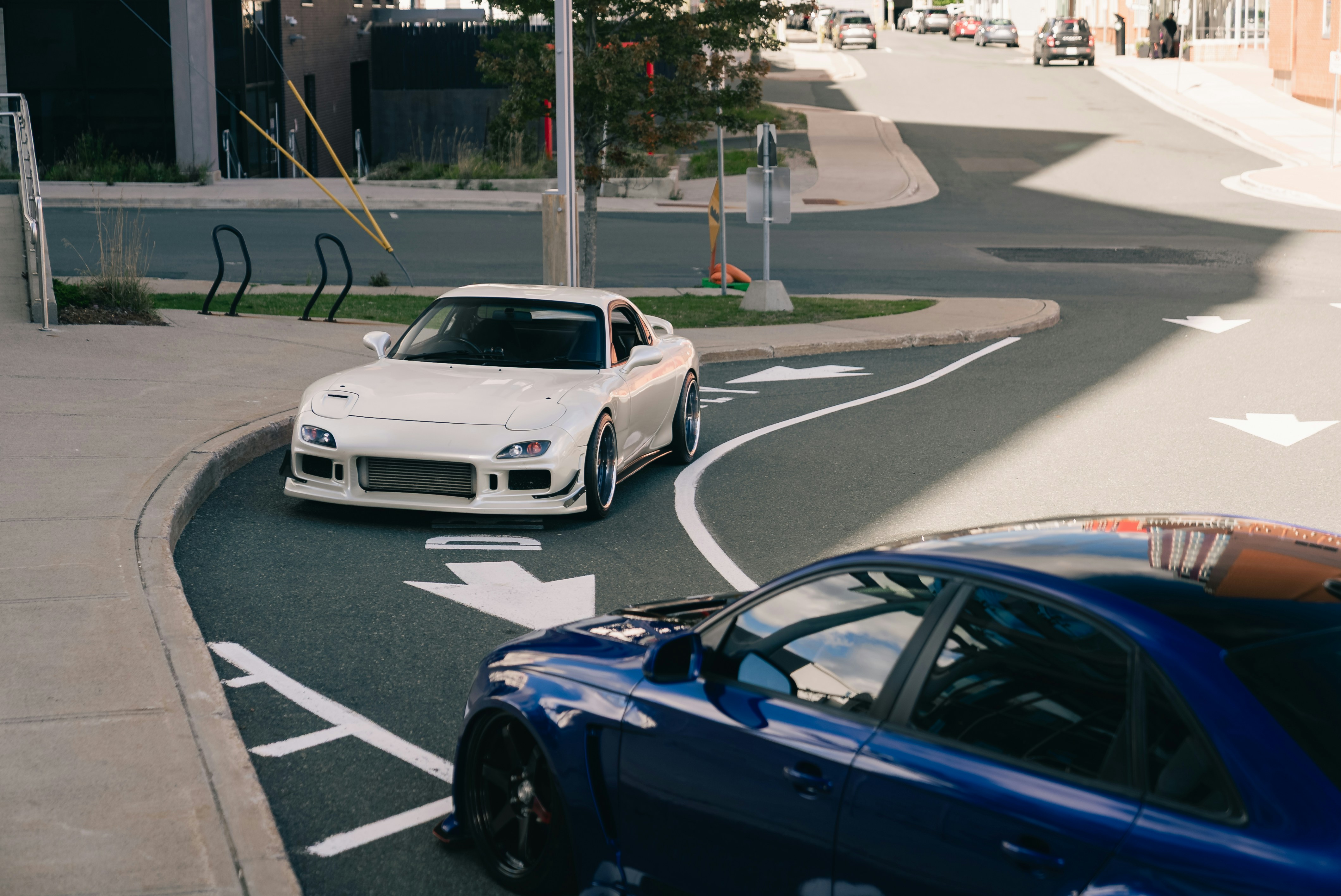 White sports car gliding past a blue coupe on a sunlit city corner