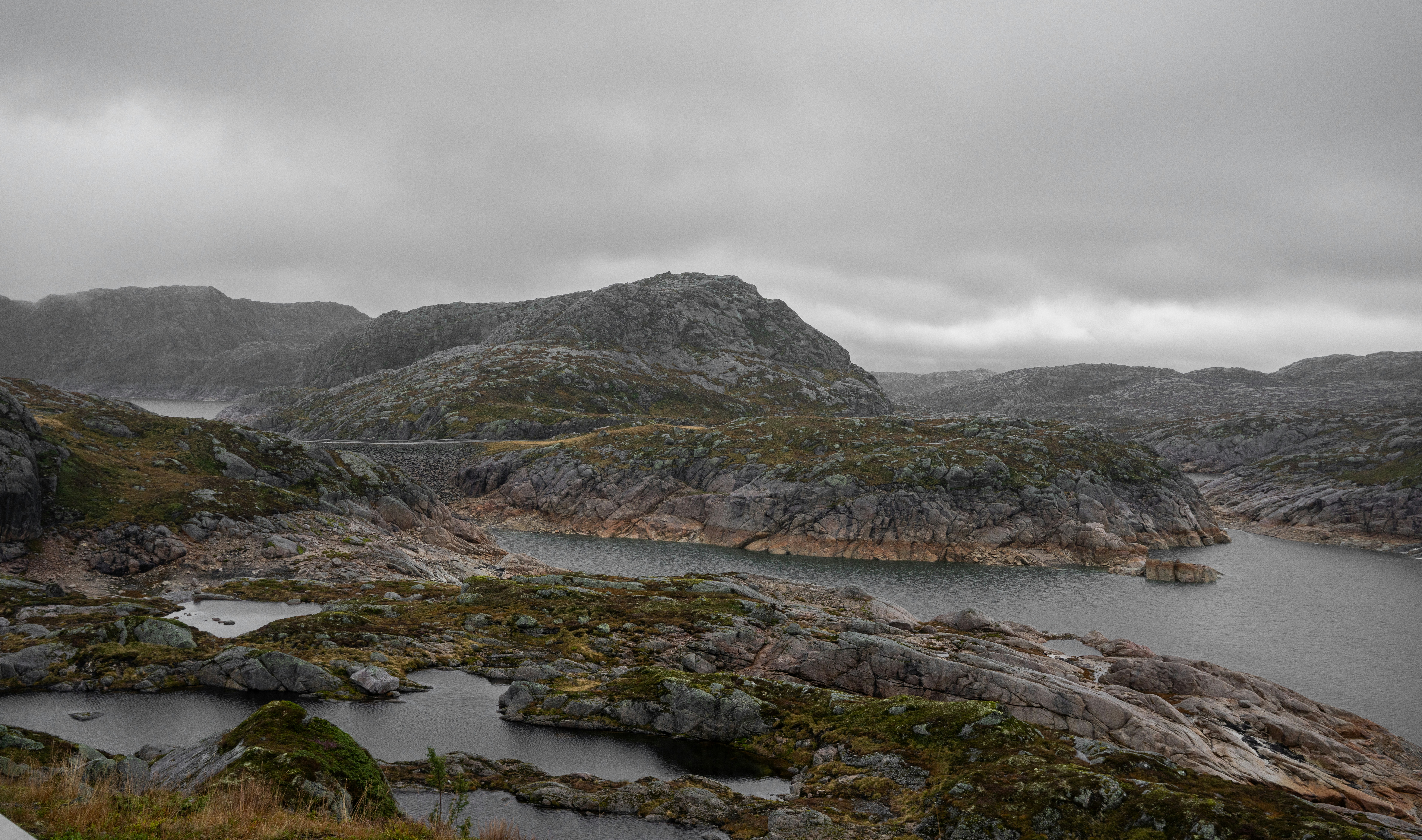 Mist-laden coastal rocks and a calm inlet framed by rugged terrain under overcast skies, portraying a remote shoreline. This is a natural landscape scene with no people or objects identifiable.