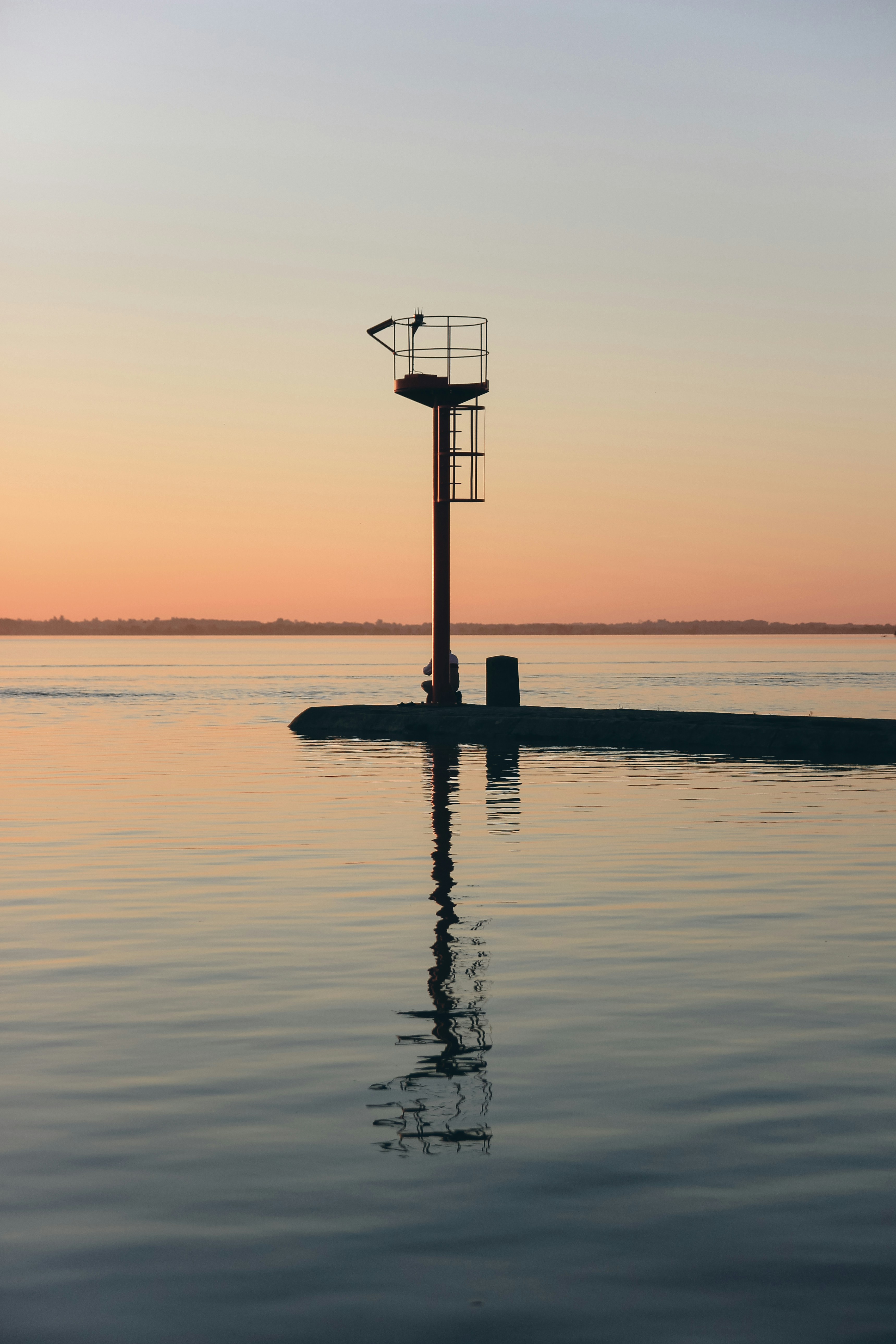 A tall navigation beacon on a pier reflecting in calm water at golden hour