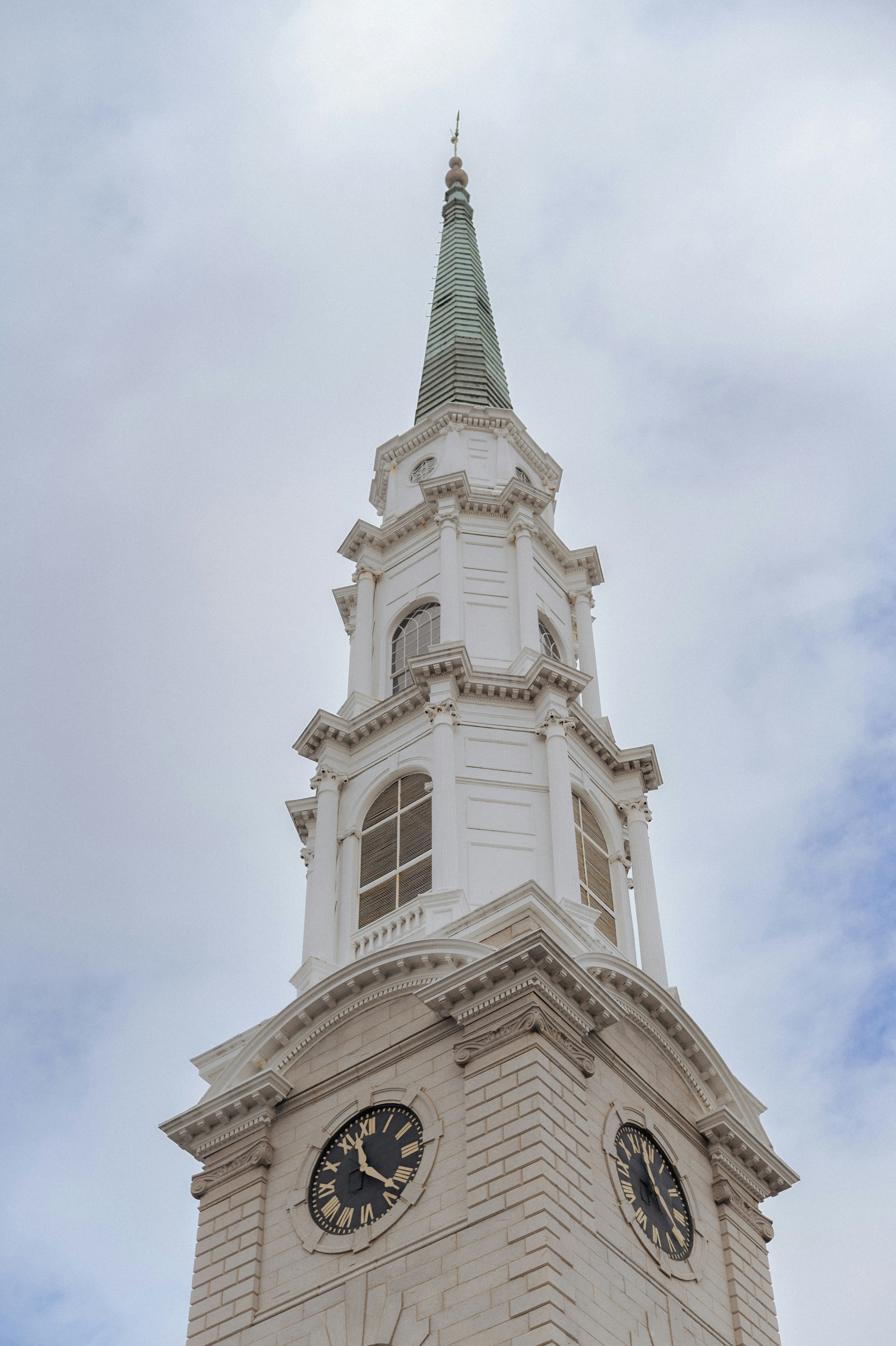 White clocktower with ornate detailing rises against a pale blue sky, the clock faces visible near the base. The structure is a historic-looking spire with multiple levels and a pointed steeple.