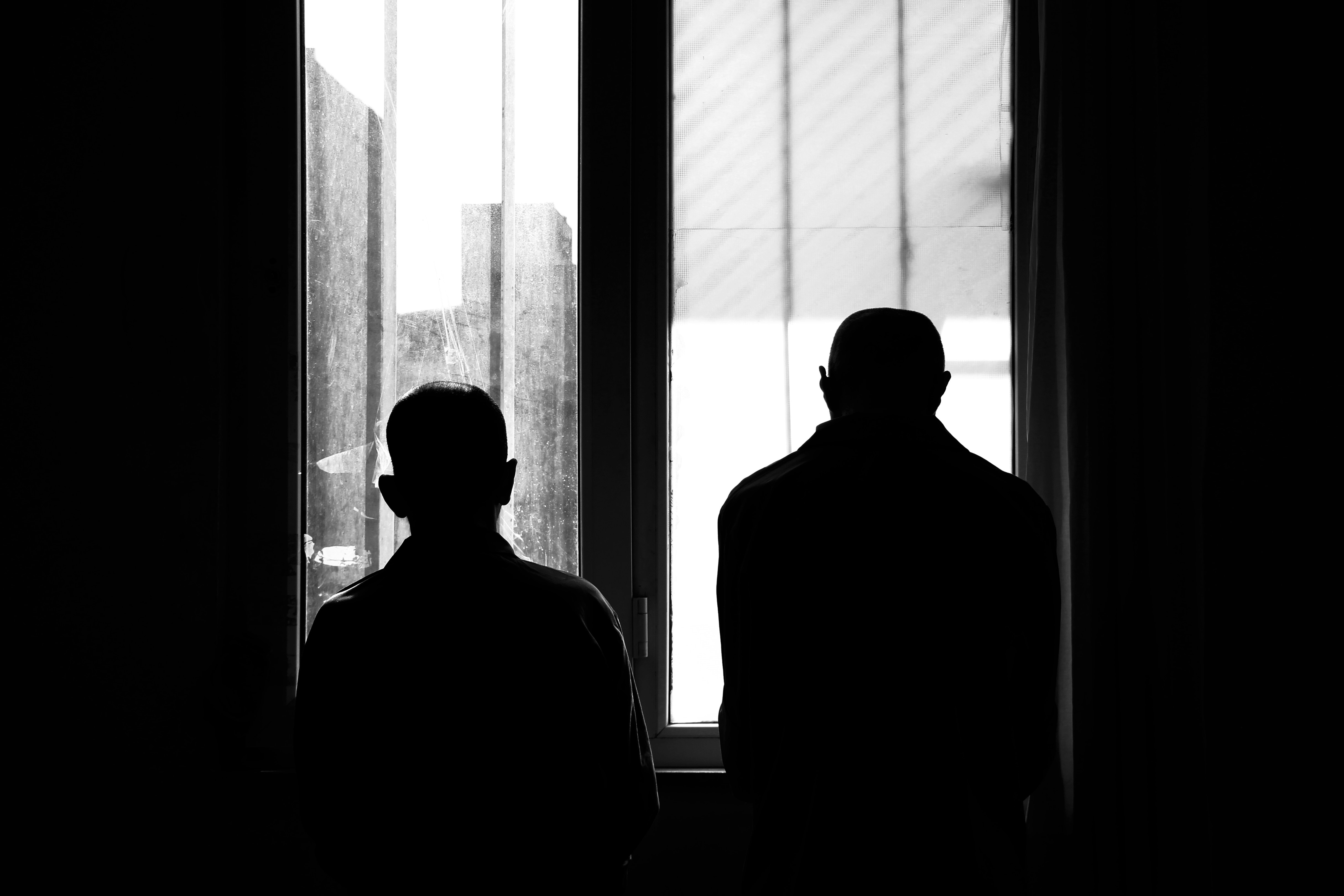 Two silhouetted figures stand before a large window, bathed in stark backlight and framed by curtains. A high-contrast, monochrome scene that emphasizes shape and negative space.