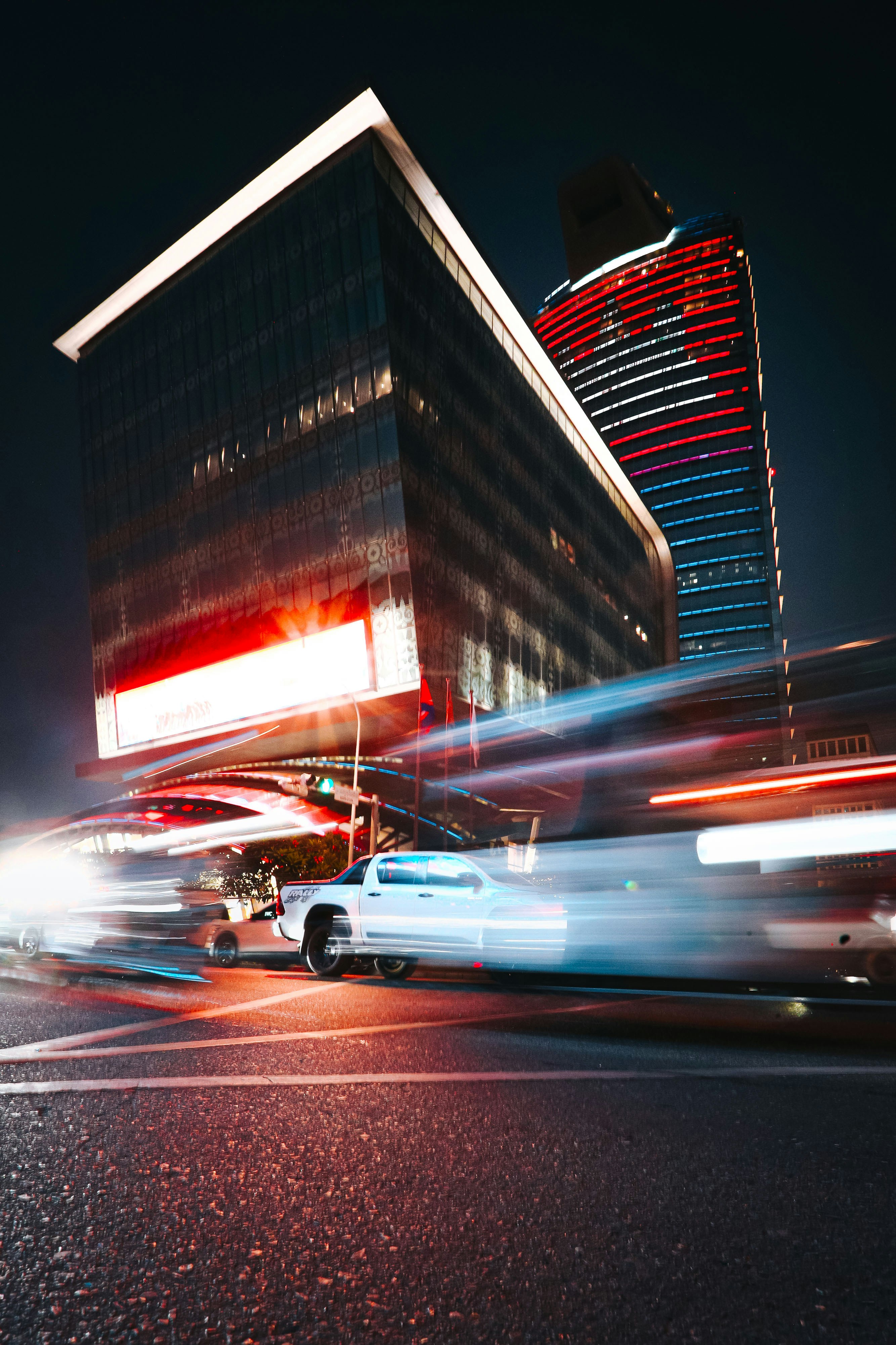 Busy urban intersection at night with motion-blur cars and a prominent glass building and illuminated tower in the background.