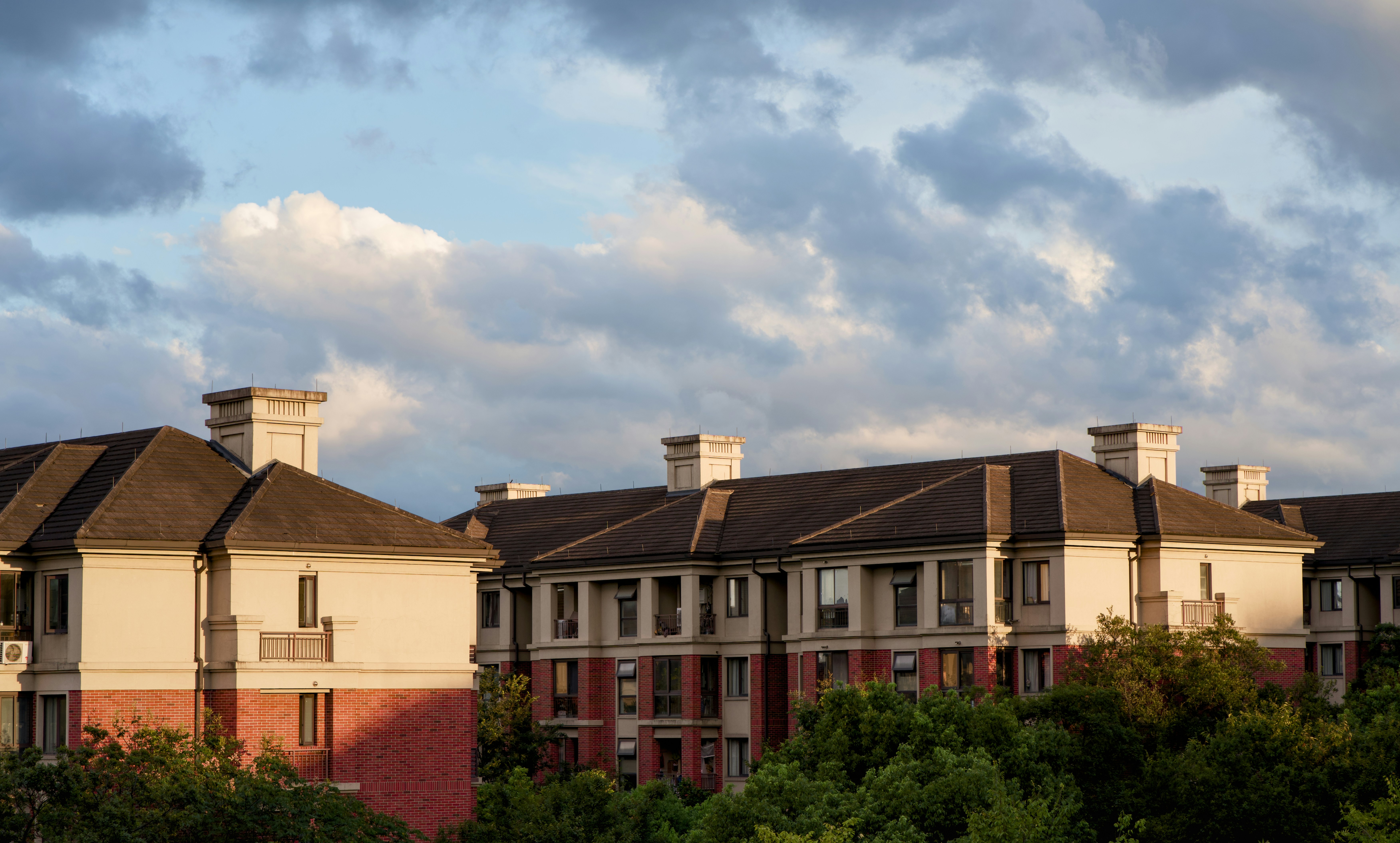 A row of campus-style buildings bathed in warm afternoon light, with green treetops in the foreground and a blue sky scattered with clouds.