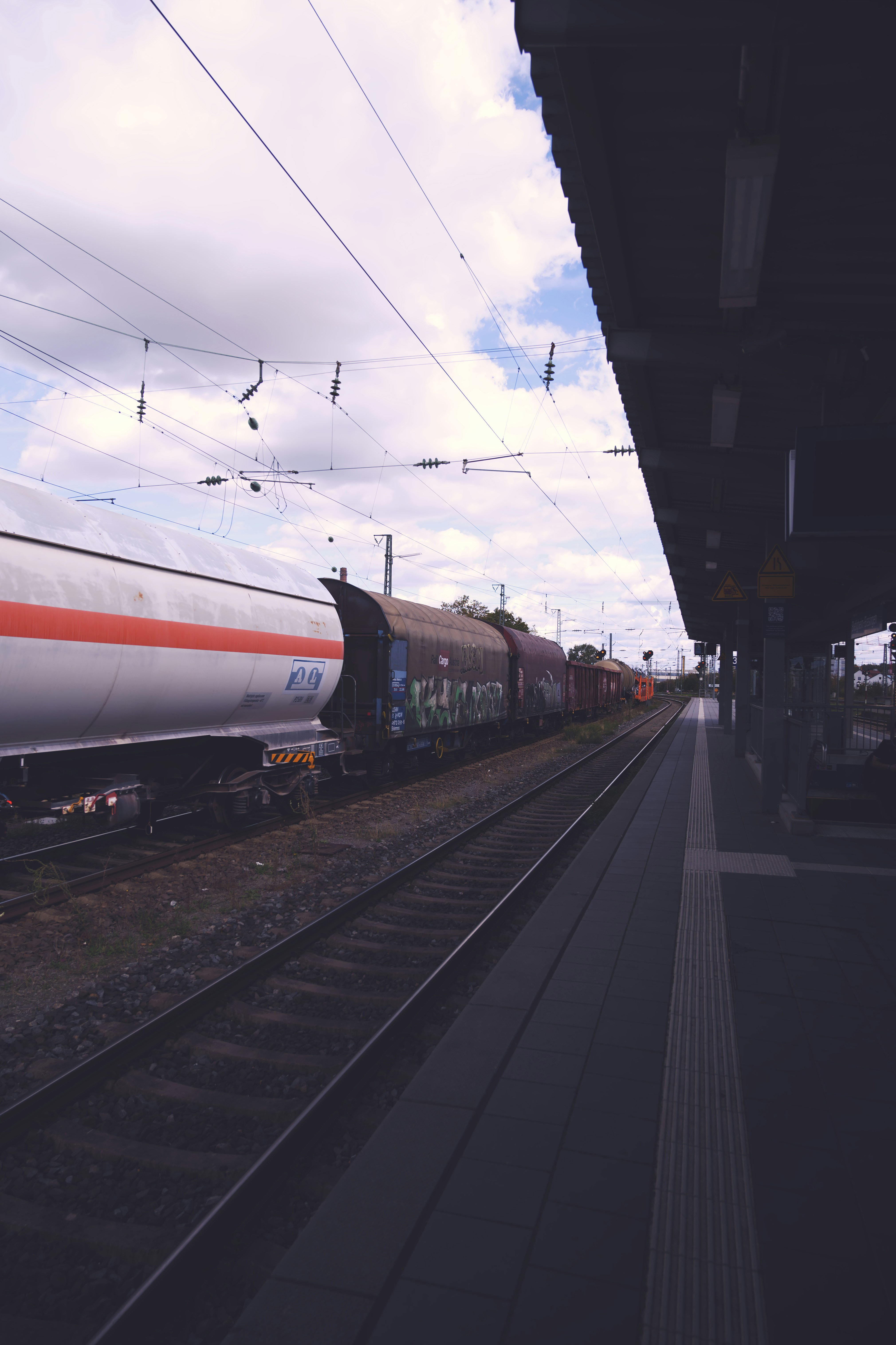 A long platform beside a freight train on tracks under a network of overhead lines at a station, with darkened shelter on the right and a moody sky. This is a photograph of a railway scene.