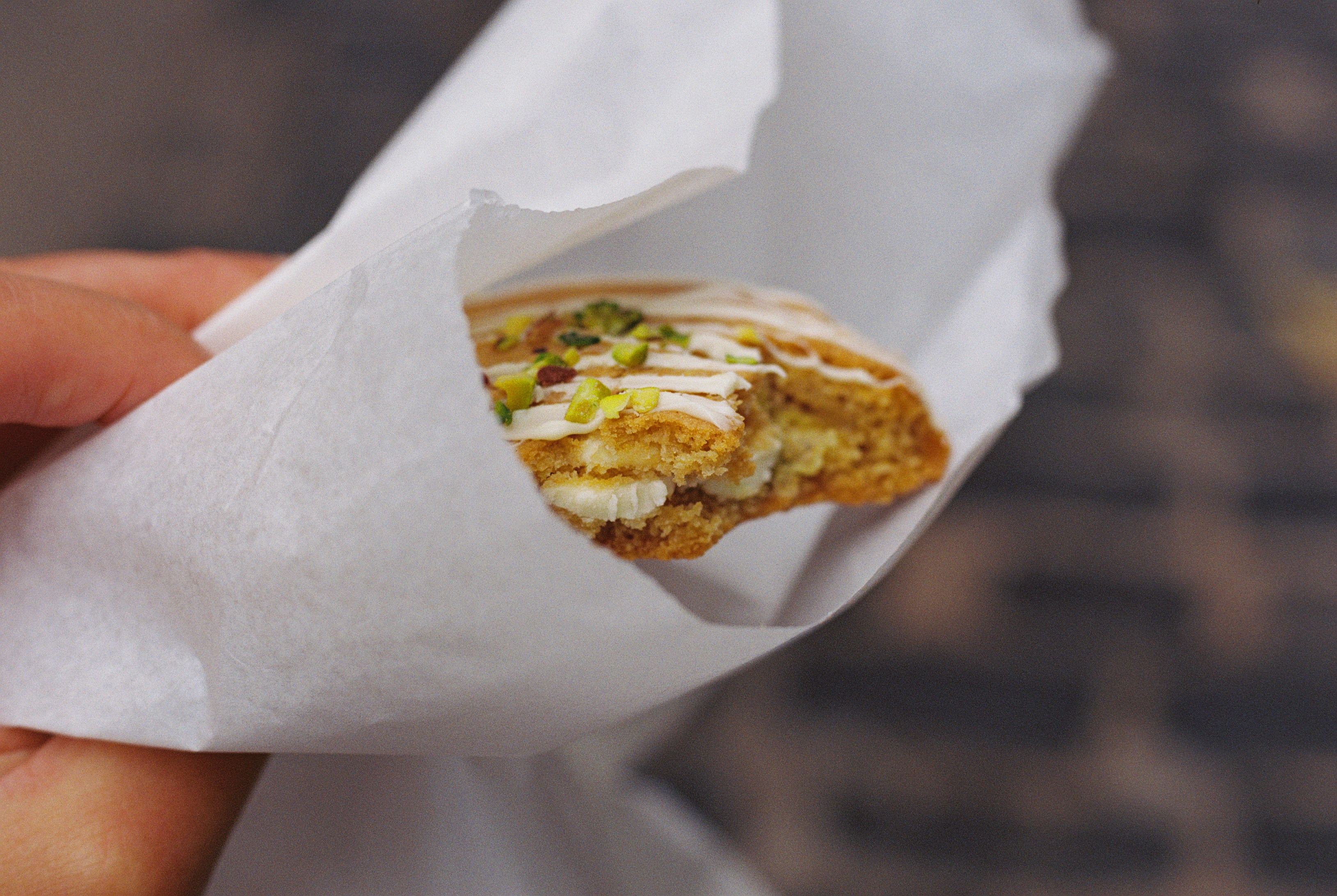 A hand holds a half-eaten iced biscuit in a twist of white paper, with the bitten edge turned towards the lens. The biscuit is golden and crumbly, striped with white icing and scattered with chopped green pistachio, and a pale filling shows in the broken section. Soft, even daylight makes the paper read as a smooth grey-white, and the dark brick-like background melts into a brown blur. Focus is tight on the bite, with a shallow depth of field and a warm yellow cast. Taken on analogue film, the picture carries fine grain and a snack-in-hand, street-food feel.