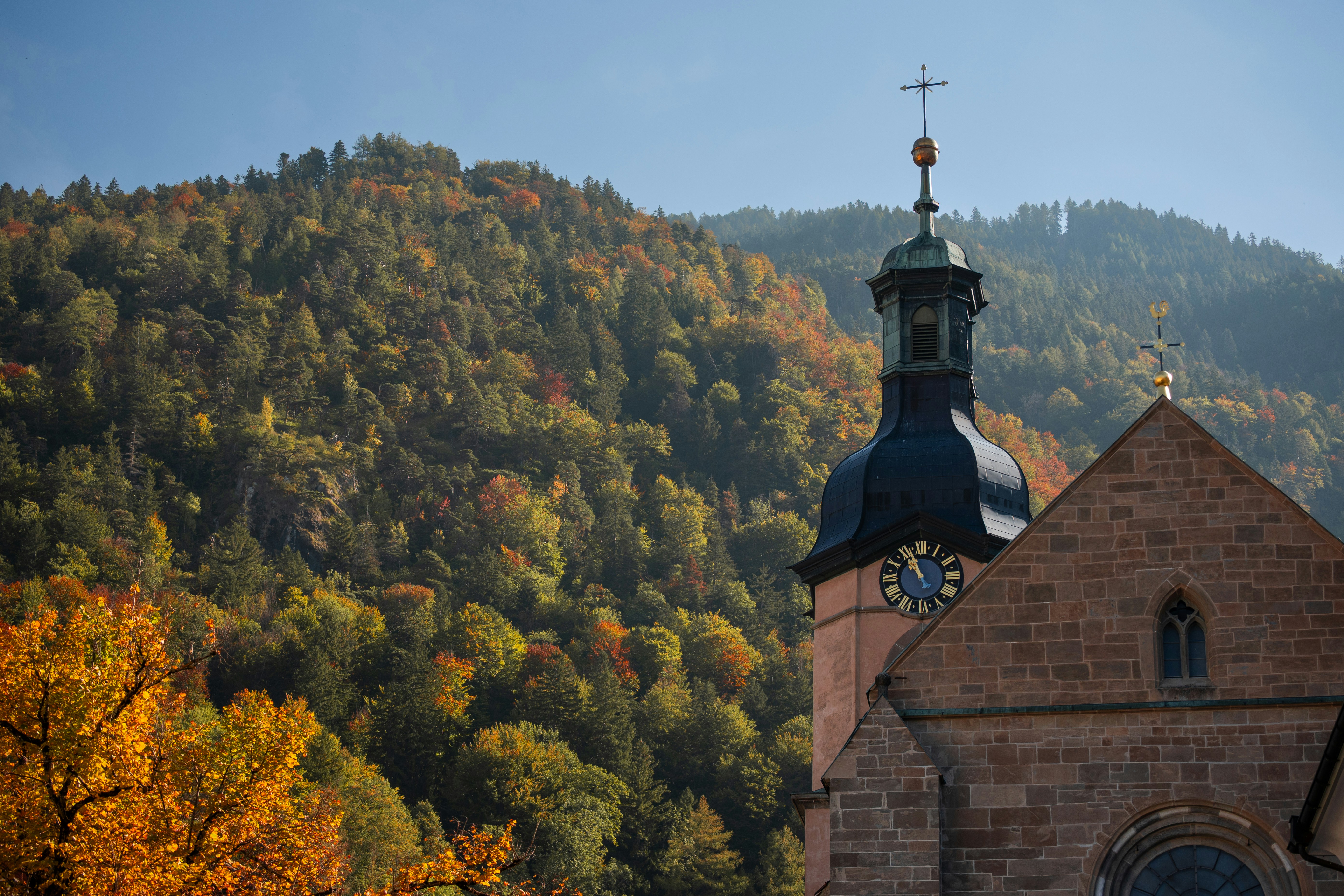 Brick church tower with clock faces rises beside a hillside forest in autumn hues, bathed in soft afternoon light.