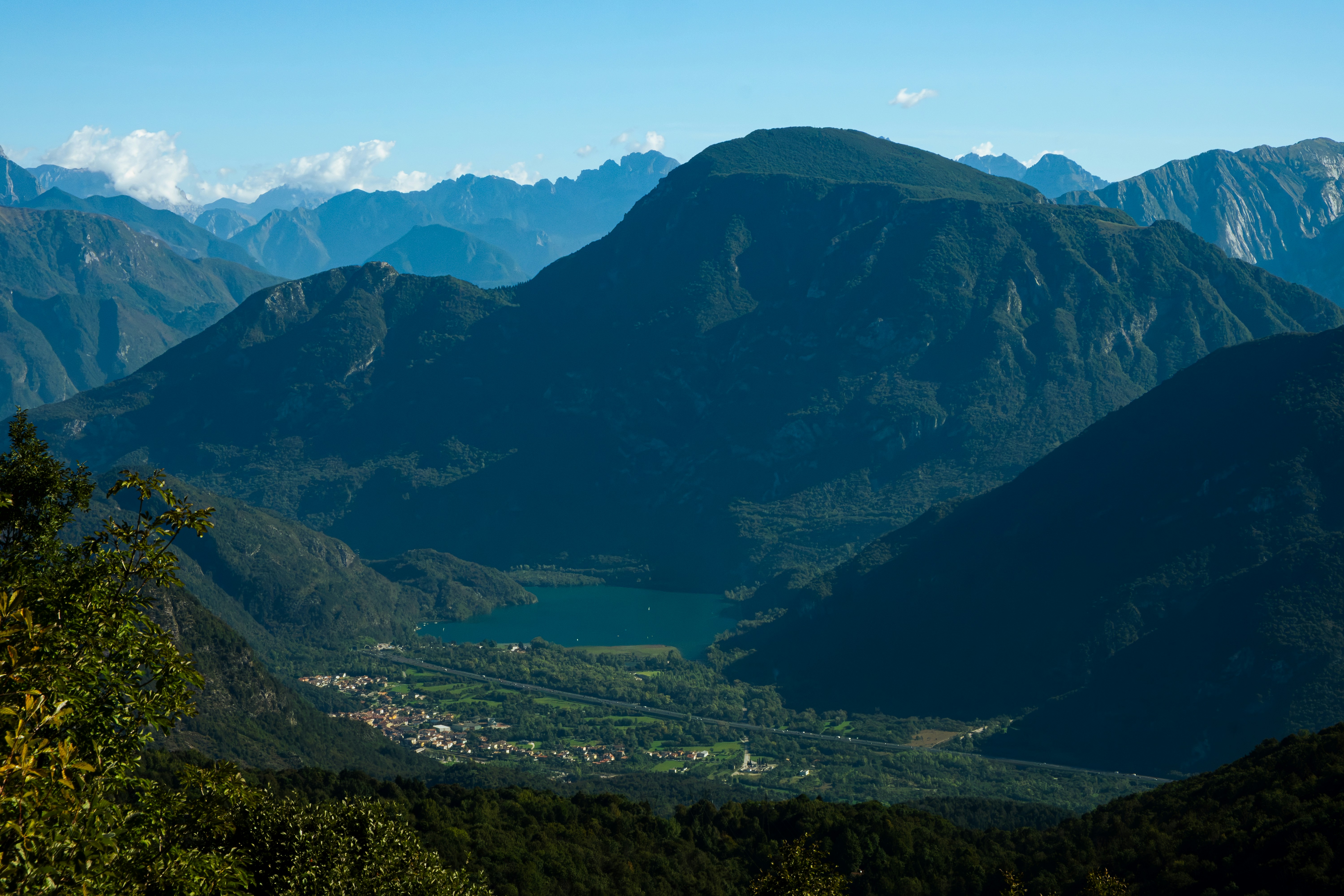 Wide valley with a lake tucked between forested mountains and a distant village, bathed in soft daylight.