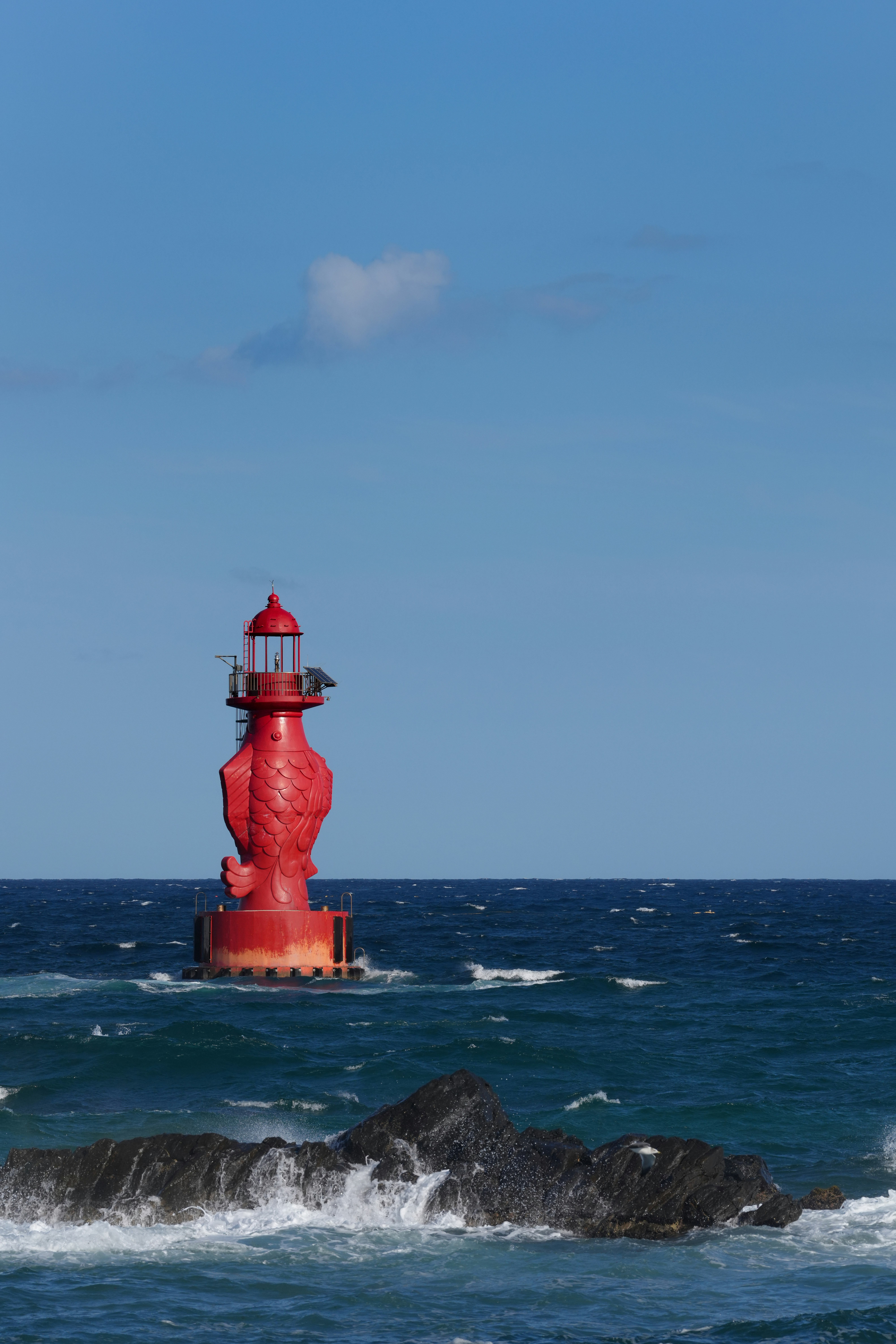 Red lighthouse statue rises from dark rocks, surrounded by ocean waves under a blue sky.