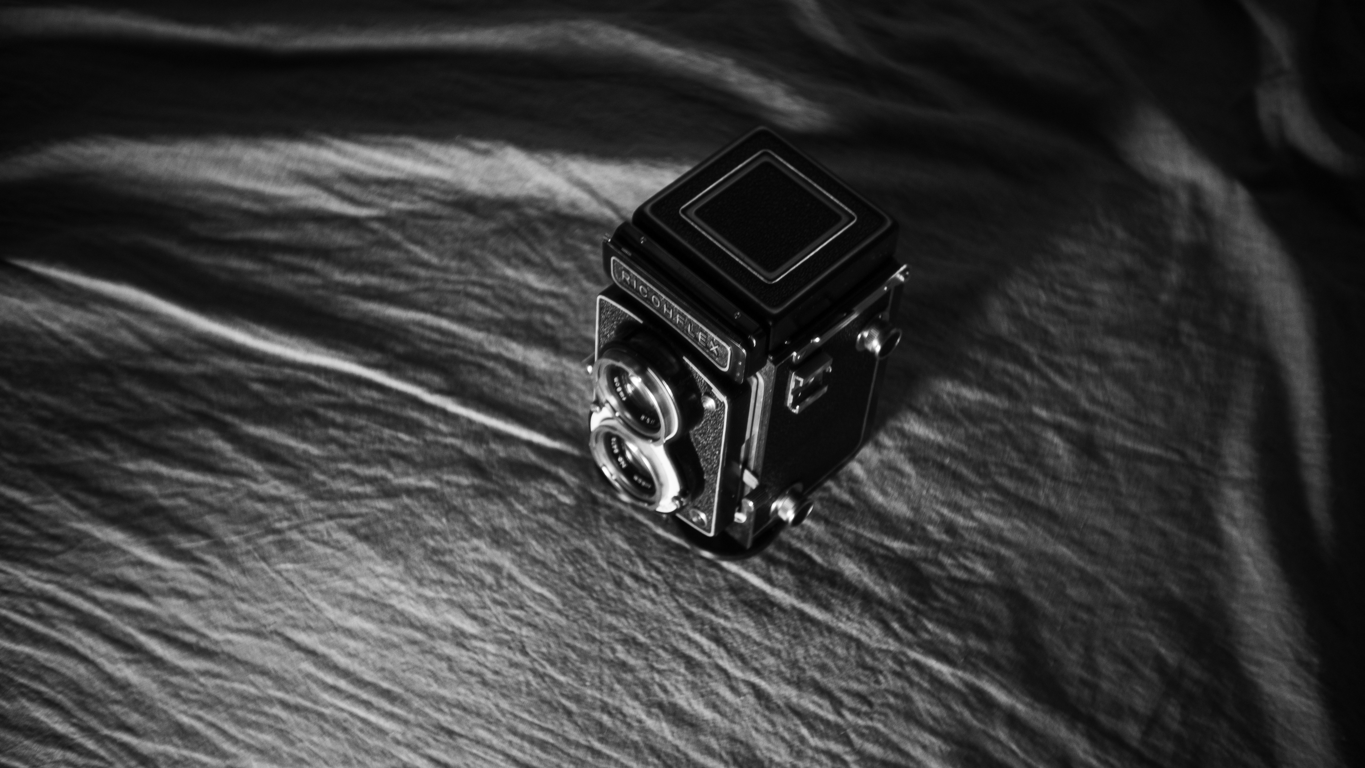 A compact vintage folding camera sits on a textured, dark fabric backdrop, captured in monochrome to emphasize its metallic details and retro design.