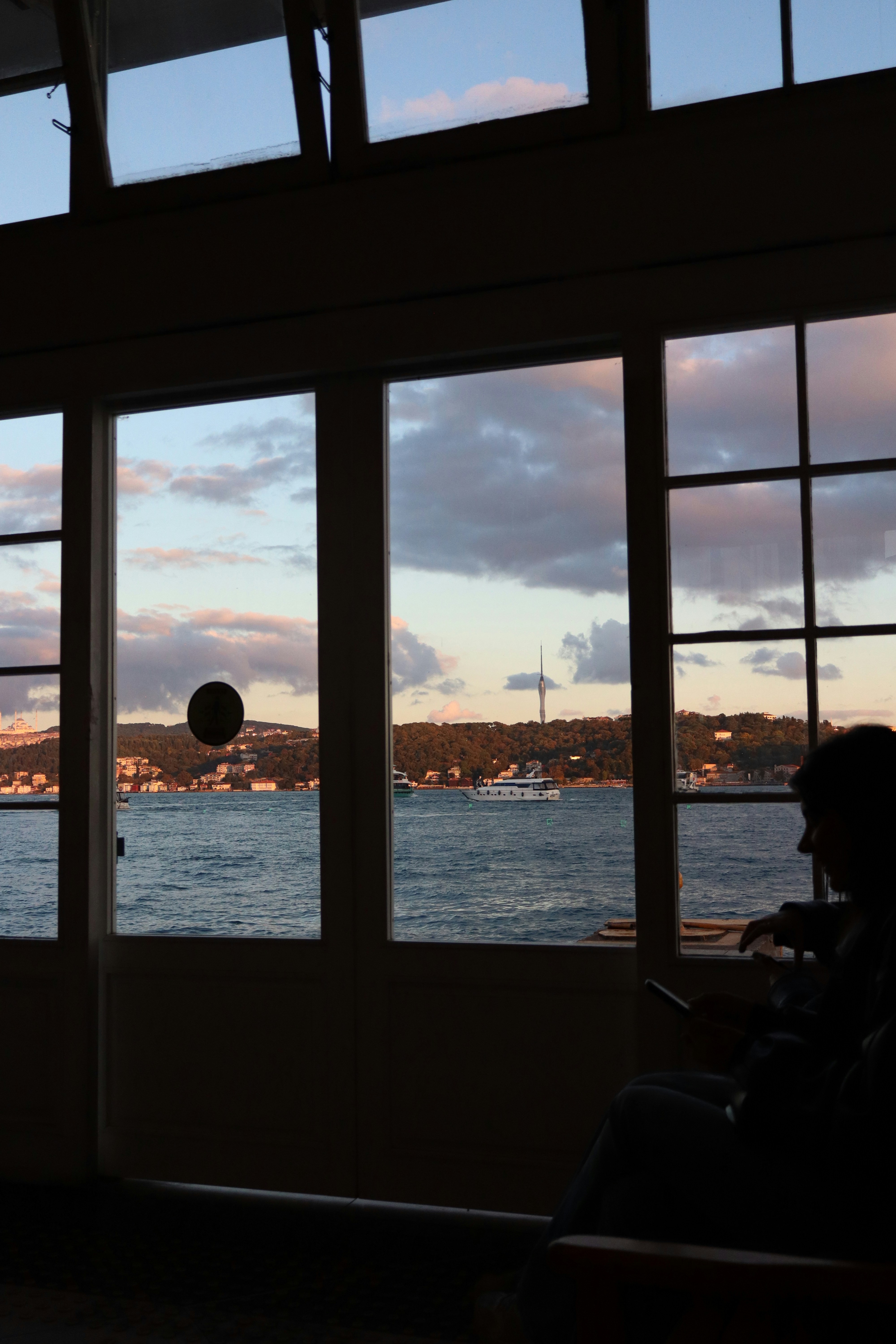 Shadowed figure by the right window looks toward a sunlit harbor with boats and distant hills visible through large panes.