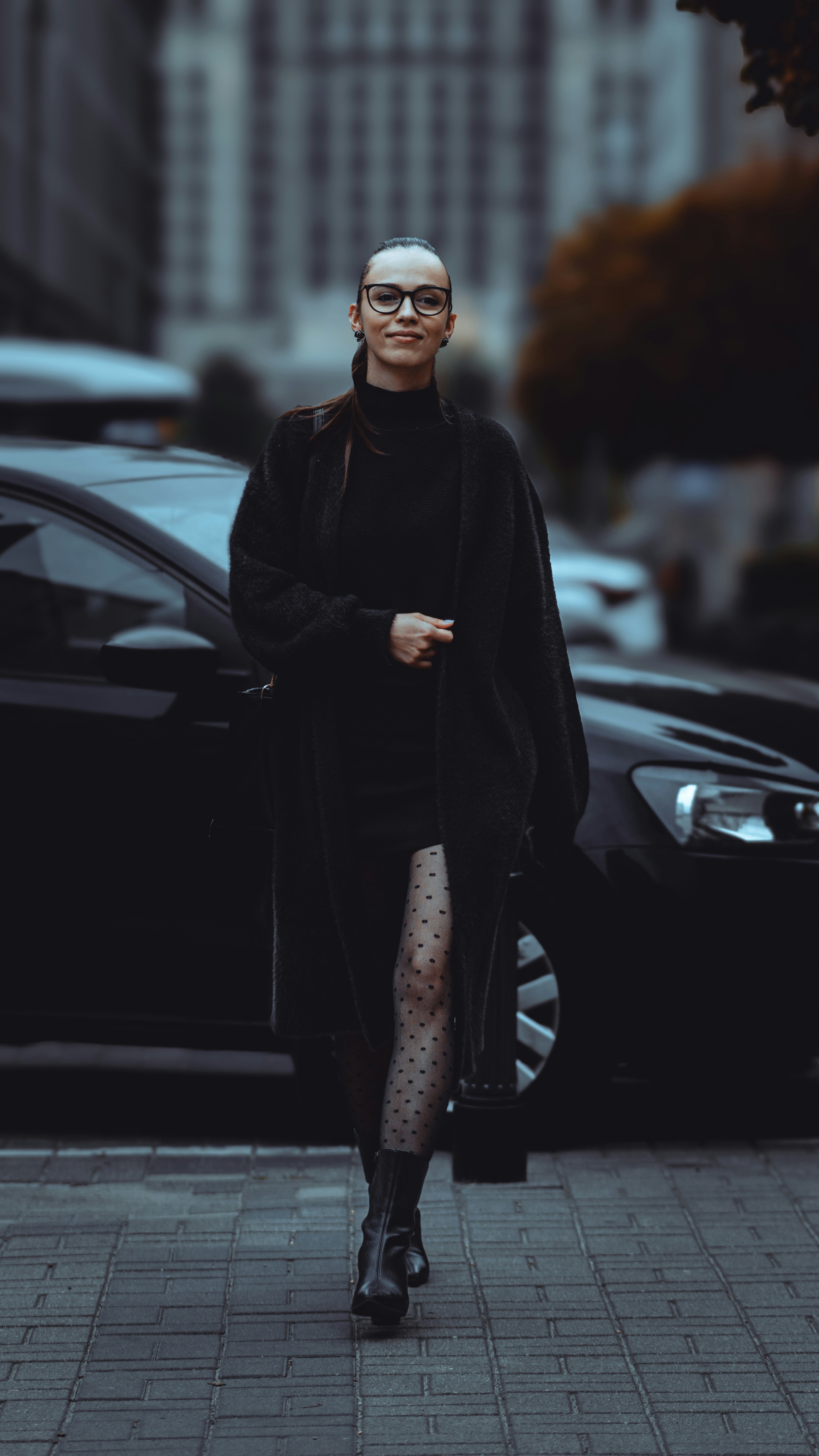 A stylish woman in a black ensemble walks past a parked car on a urban street, blurred cityscape behind. The image is a fashion portrait with a moody, cinematic tone.