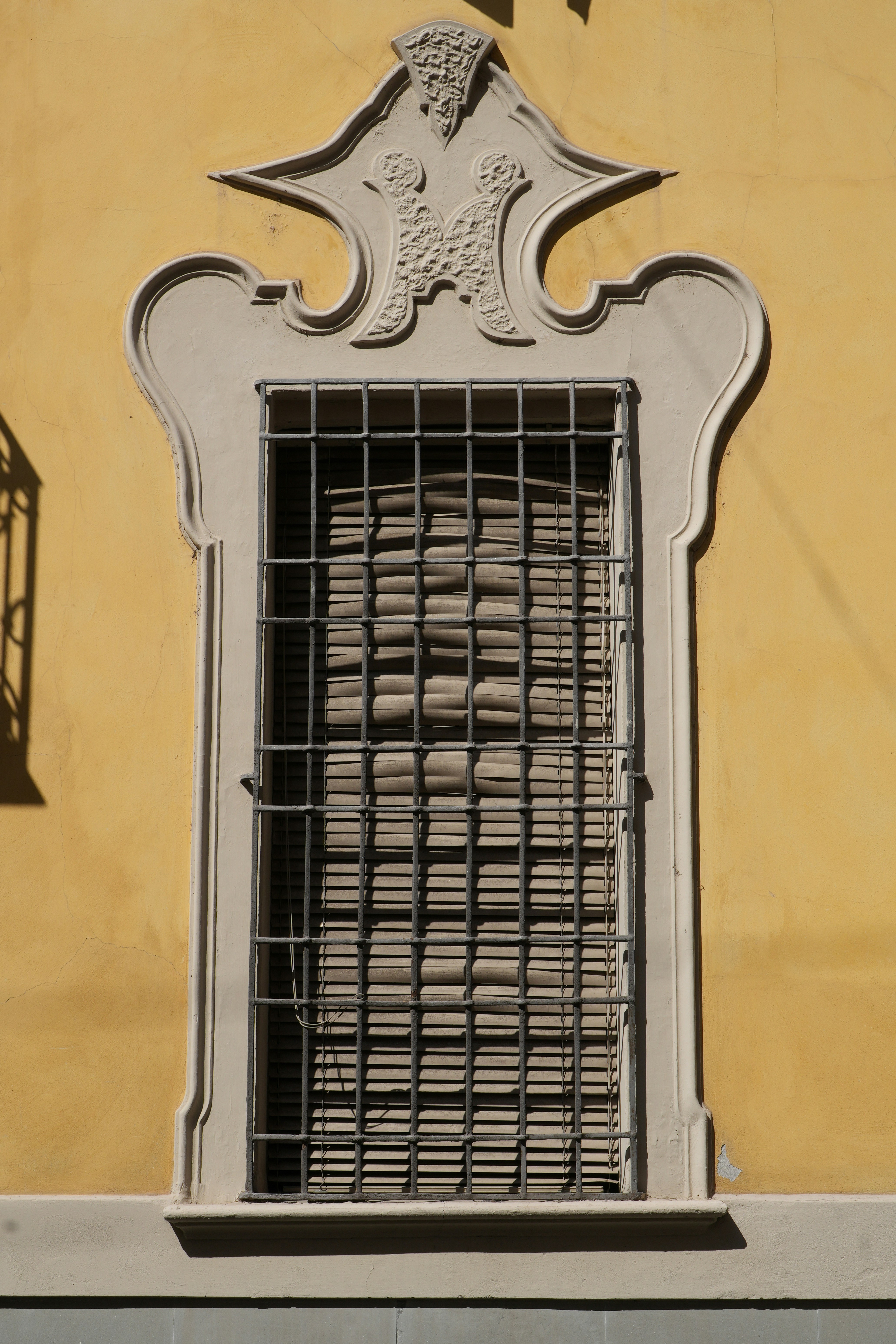 Bright yellow wall with an elaborately shaped window opening secured by a metal grille; the shuttered interior shows layered slats, set on a stone sill.