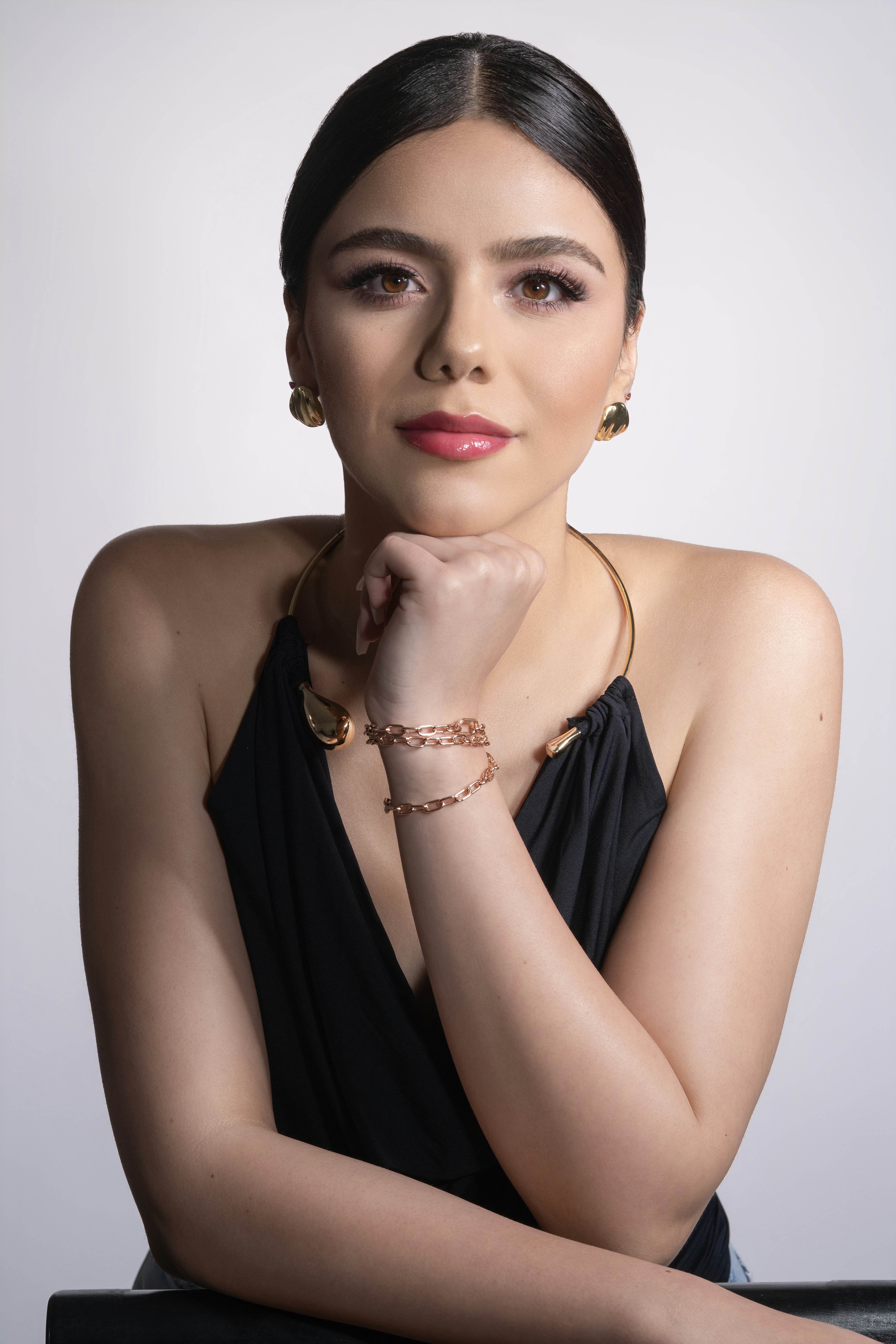A close-up portrait of a poised woman with dark hair, wearing gold jewelry, resting her chin on her fist against a neutral backdrop. The image is a studio portrait with a wardrobe accent and soft lighting.