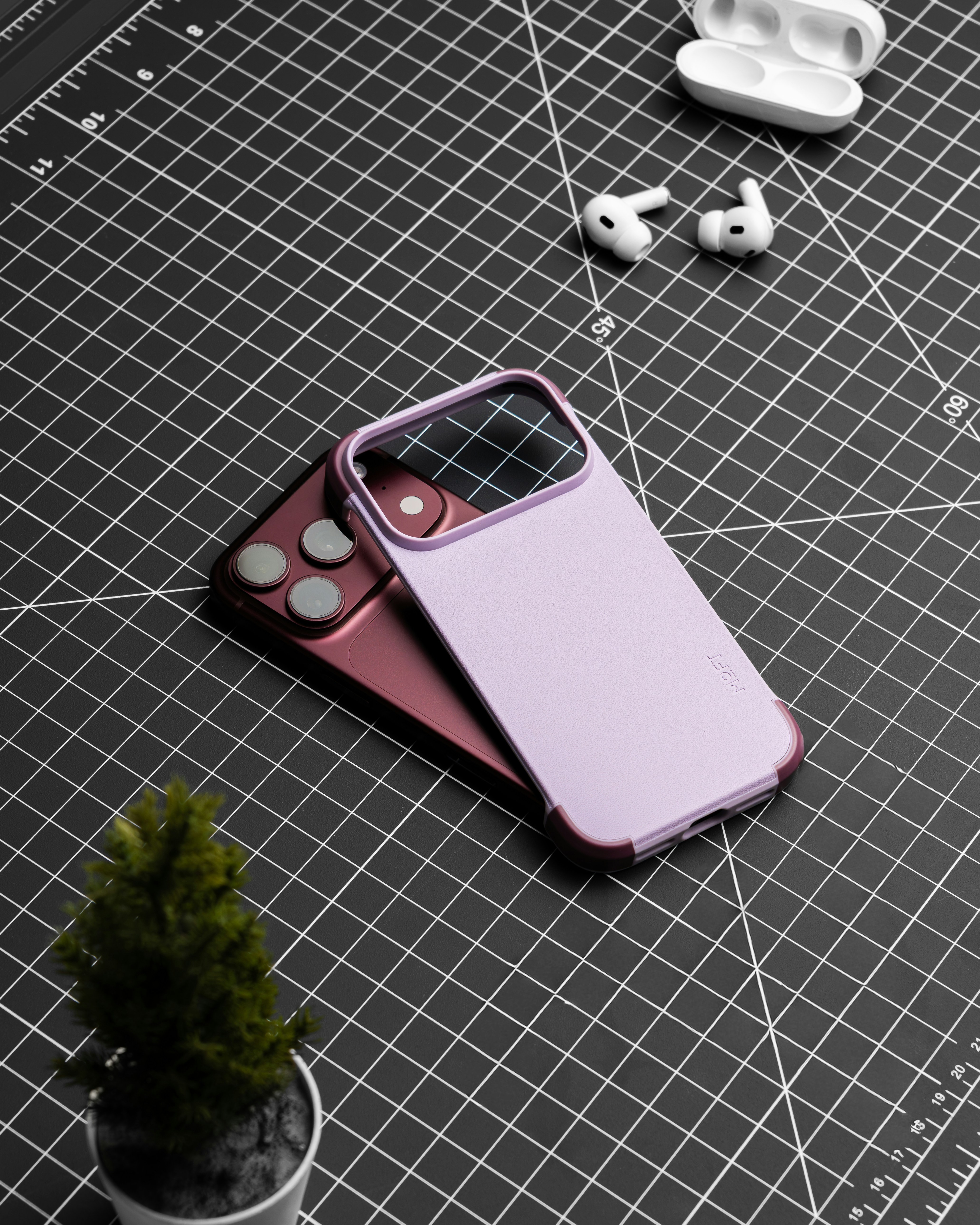 Compact tech setup featuring a lavender phone case placed on a rose-colored iPhone, accessory earbuds in the background, and a tiny potted plant on a grid cutting mat.