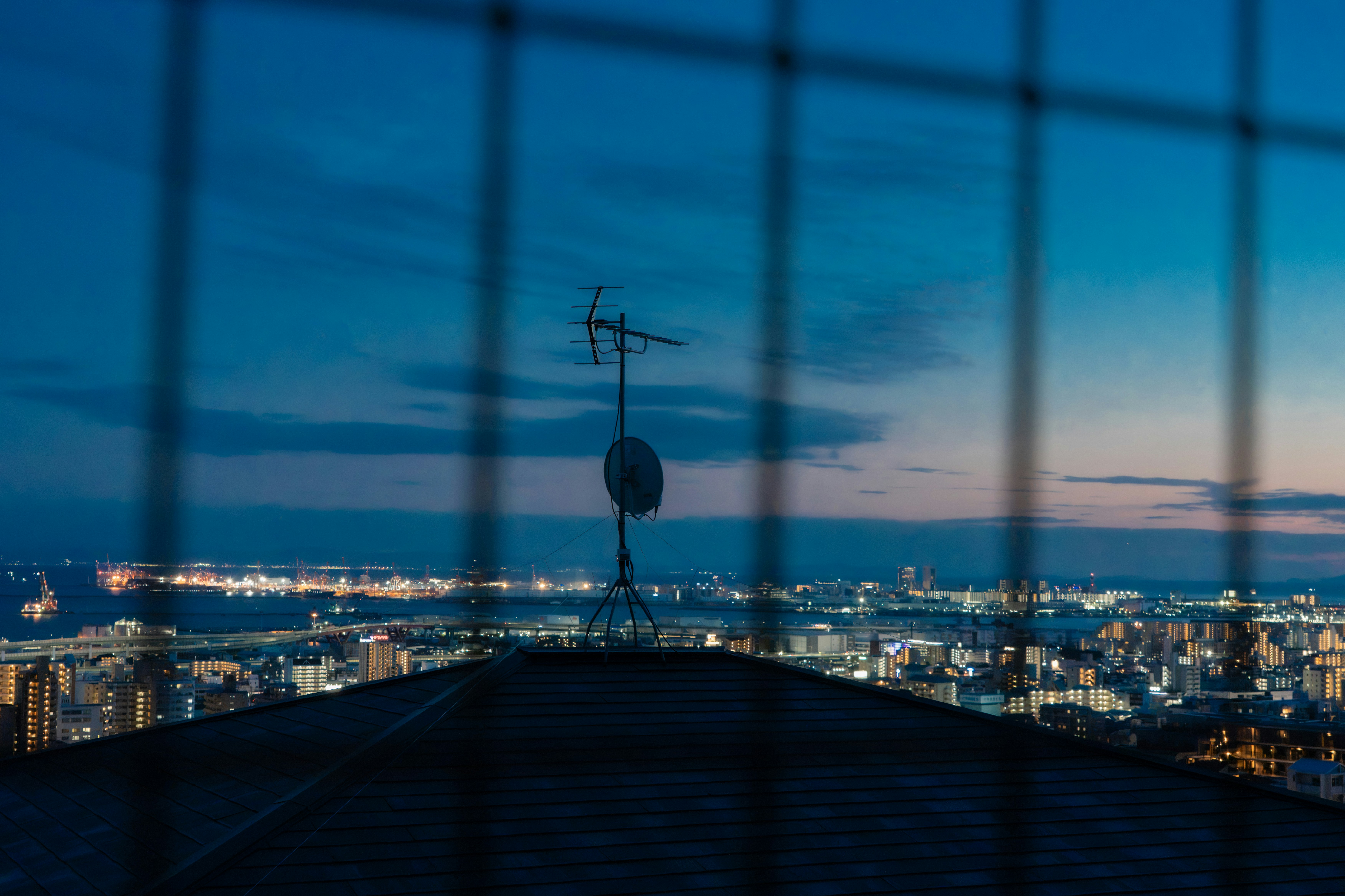 City skyline at dusk seen from a rooftop with a silhouetted antenna; soft blue twilight over illuminated buildings in the distance.