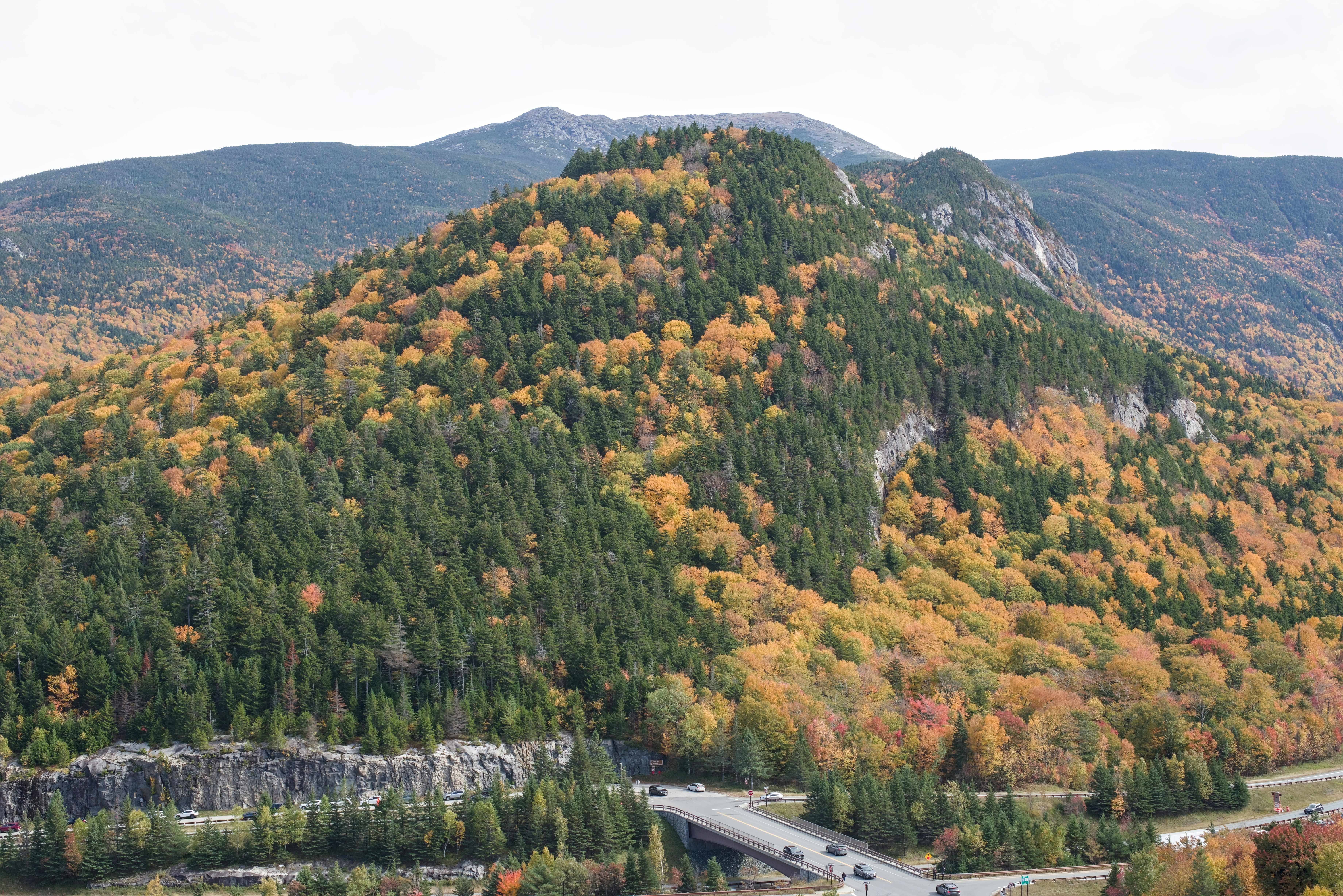 Majestic autumn-covered mountains dominate the frame, with a winding road cut below and a mix of green pines and gold-leaved trees showcasing seasonal transformation.