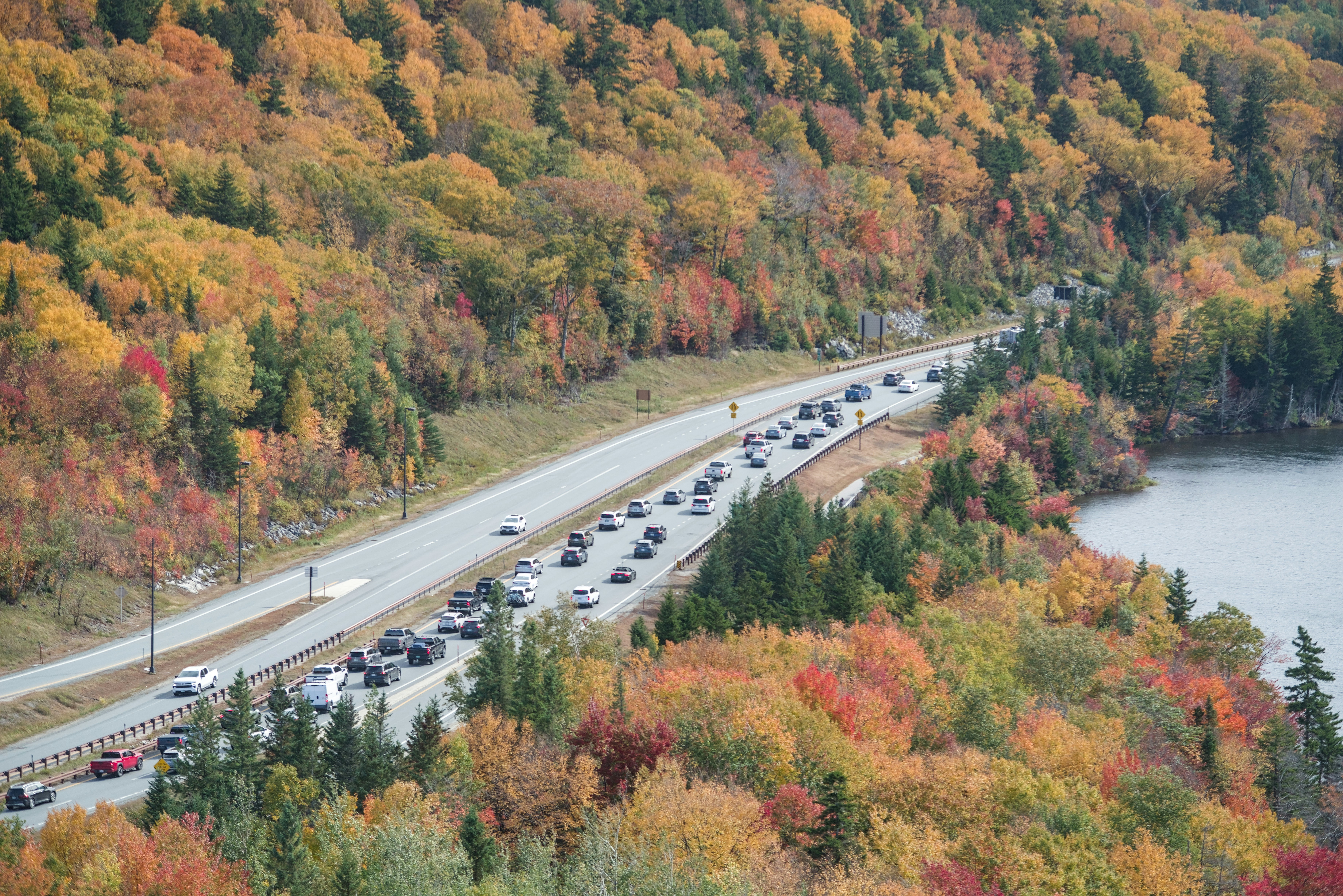 Vehicles stream along a winding highway bordered by vibrant fall foliage and a lakeside edge.