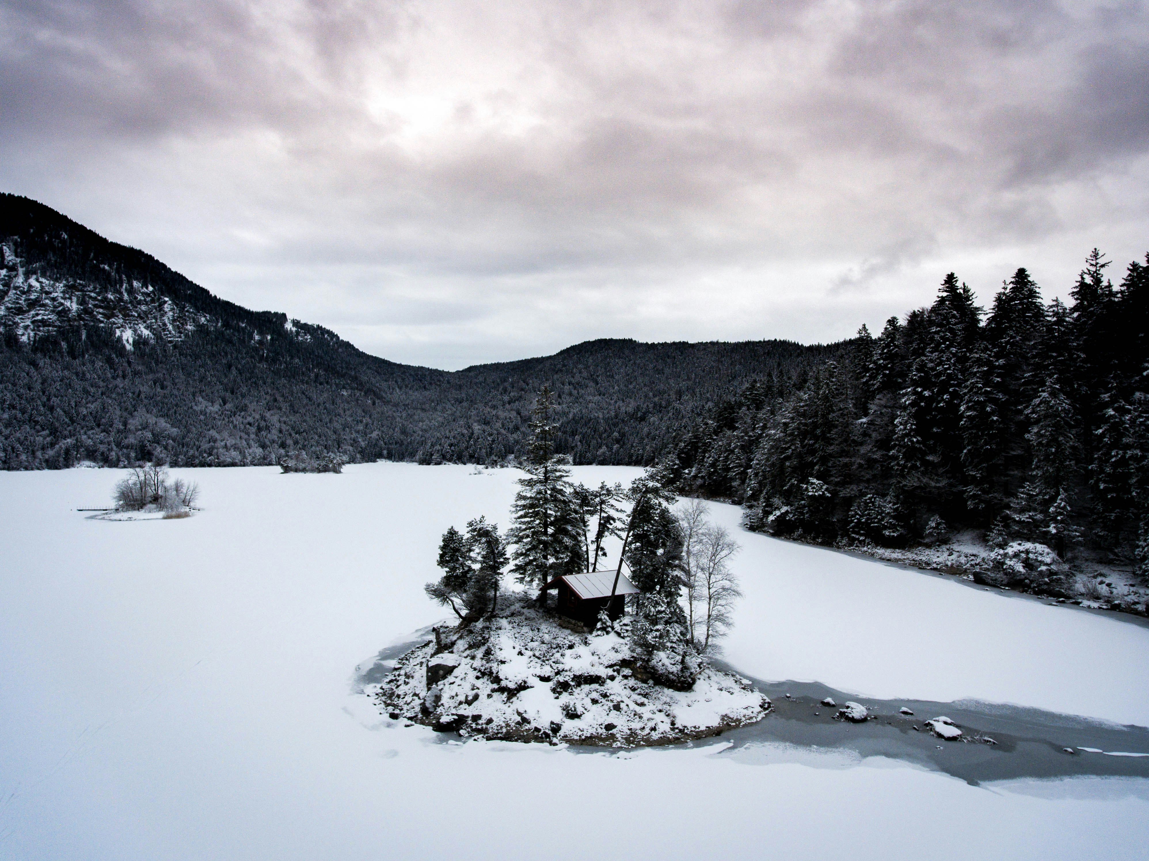 Snow covers a tranquil lake, with a small island featuring a rustic shelter and surrounding evergreen trees; distant mountains form a moody, wintry backdrop.