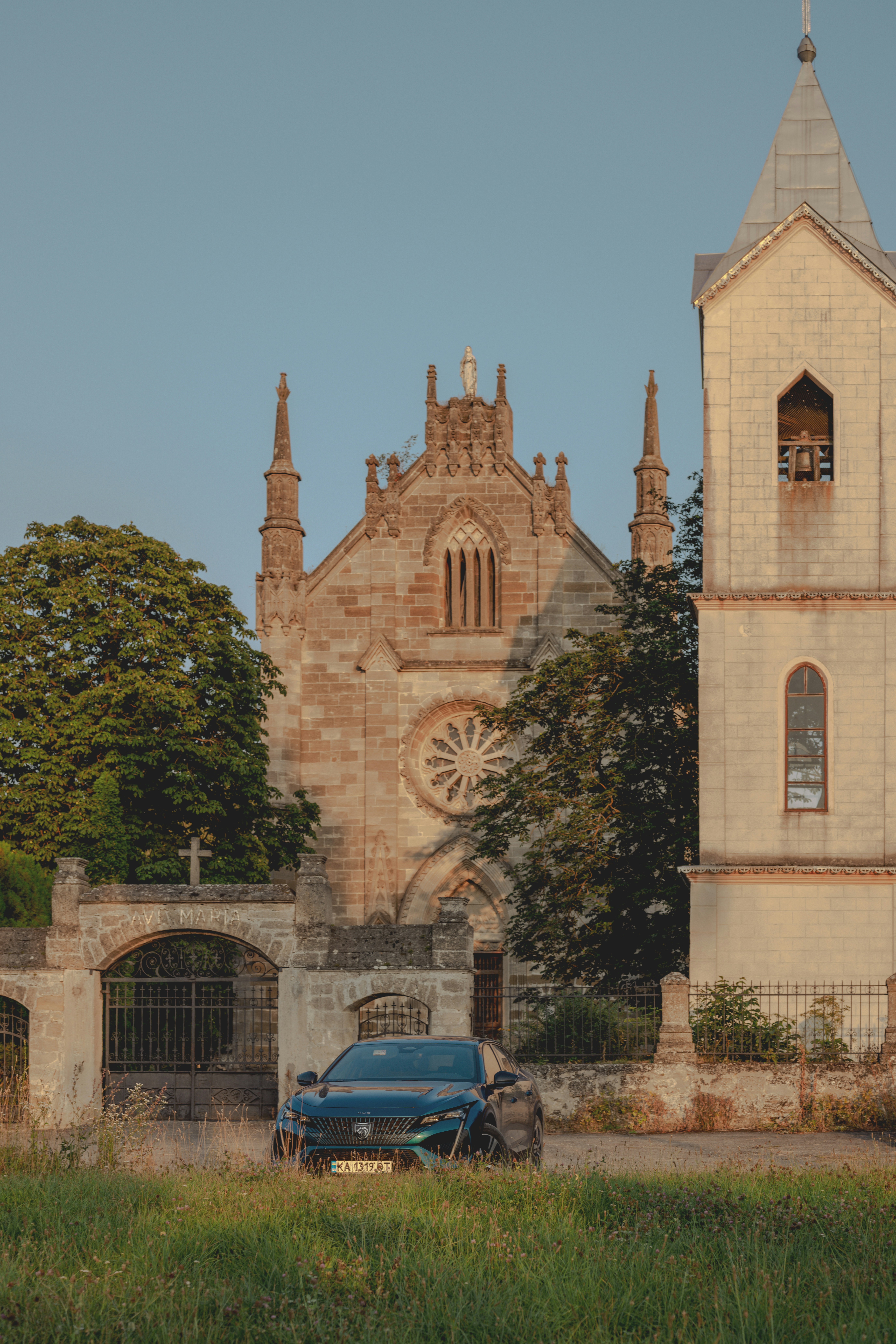 A blue car parked in front of a historic church with a tall bell tower on a grassy foreground during golden hour.