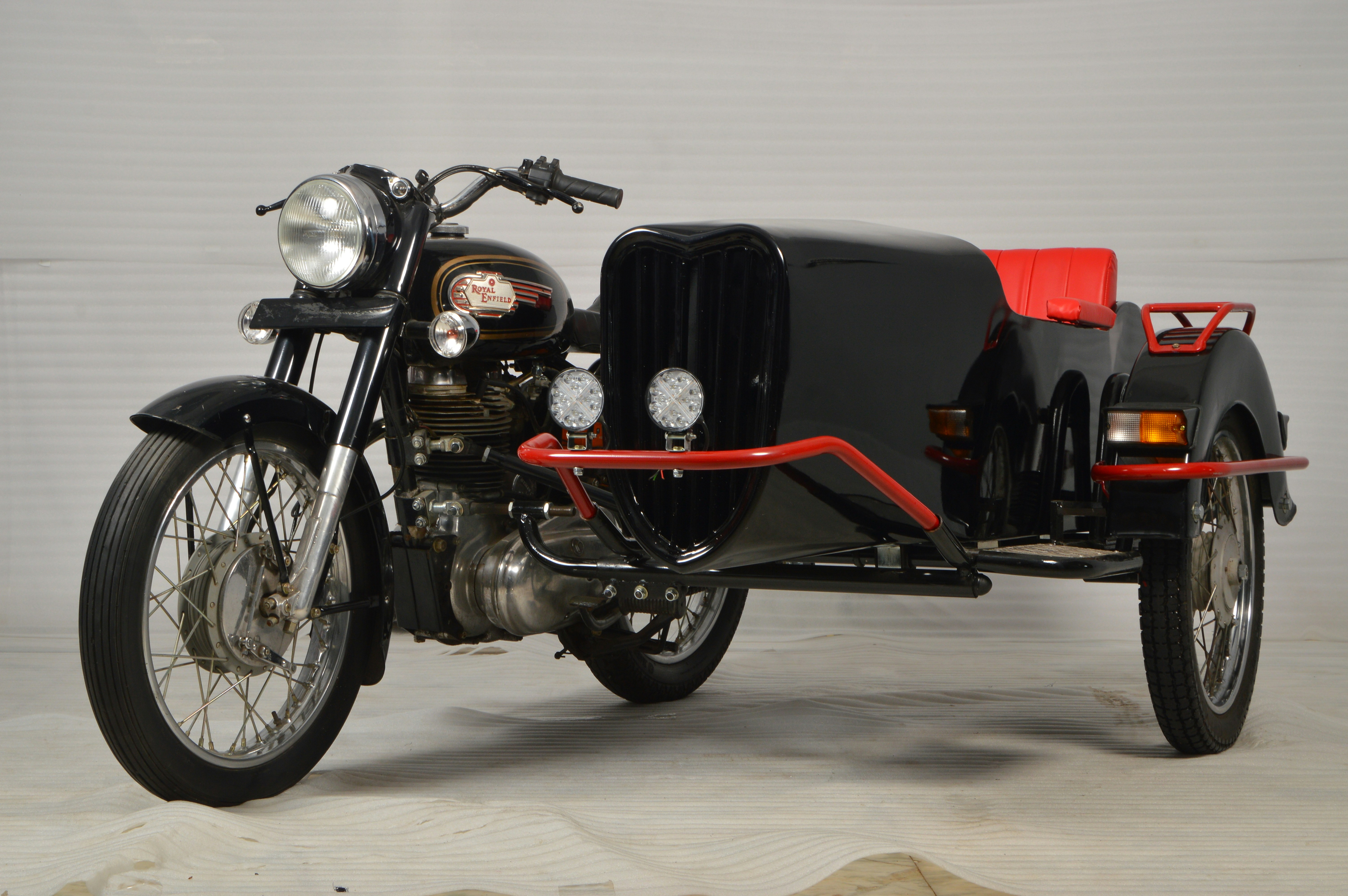 Vintage motorcycle with a red-accented sidecar frame and exposed engine, posed in a studio with neutral background to highlight its chrome and black finish.