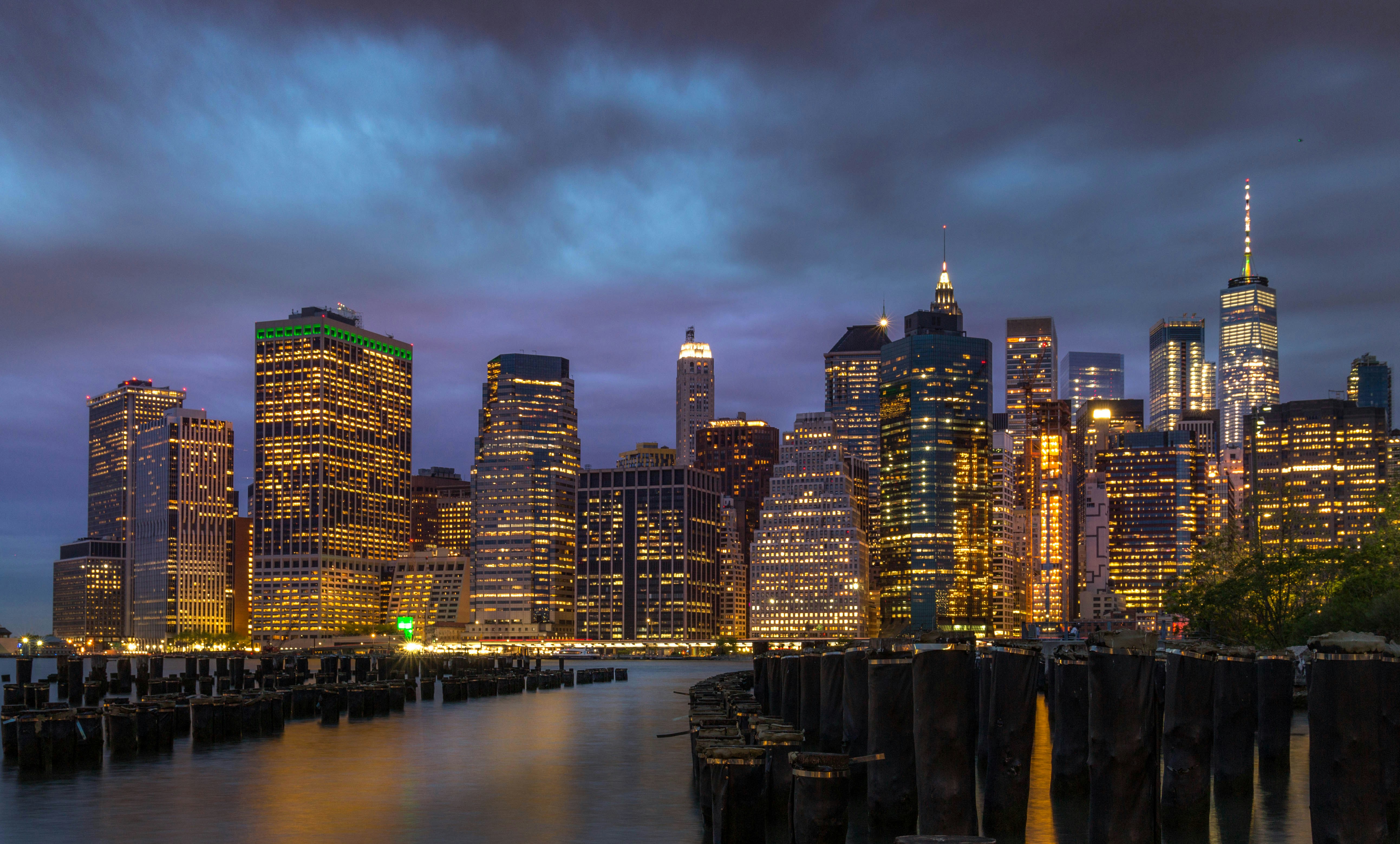 City skyline at twilight over a calm harbor with illuminated skyscrapers and wooden pilings in the foreground.