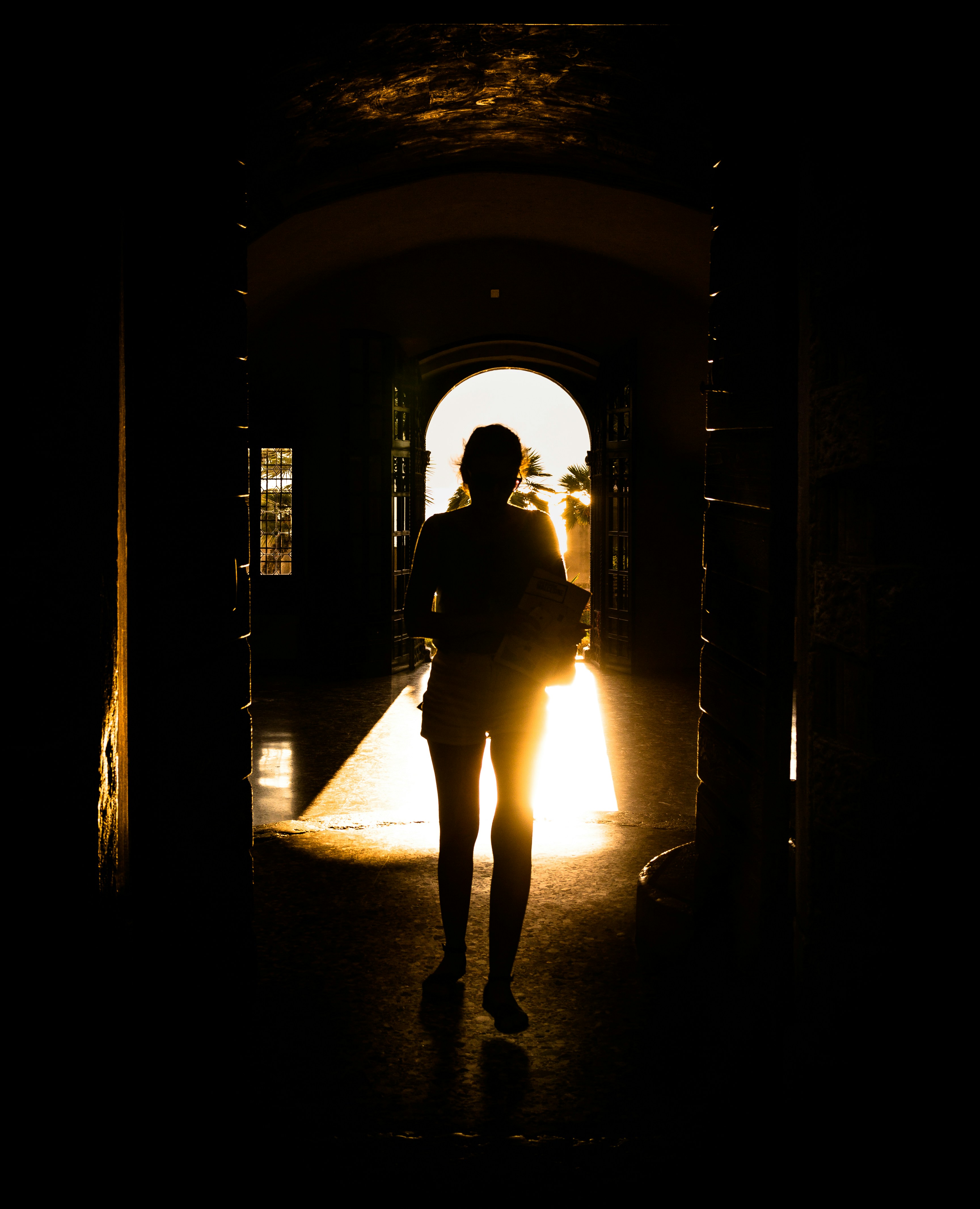 A lone person walks through a dark historic passageway toward a brightly lit doorway, creating a dramatic silhouette. A moody architectural photograph emphasizing light and shadow, perspective, and the atmosphere of historic Italian spaces.