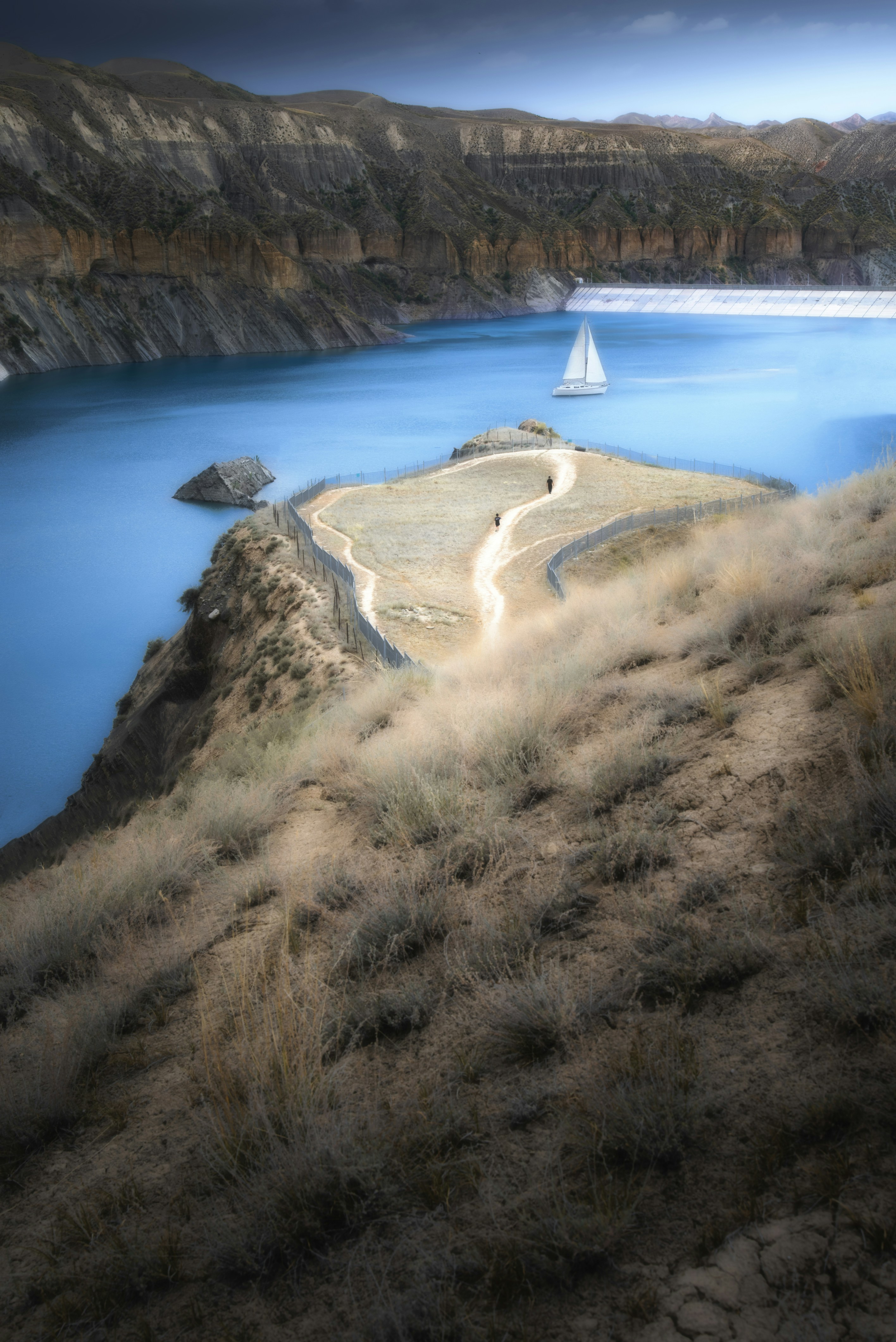A narrow peninsula curves into a vibrant blue reservoir, with a solitary sailboat near the horizon and rugged cliffs framing the scene.