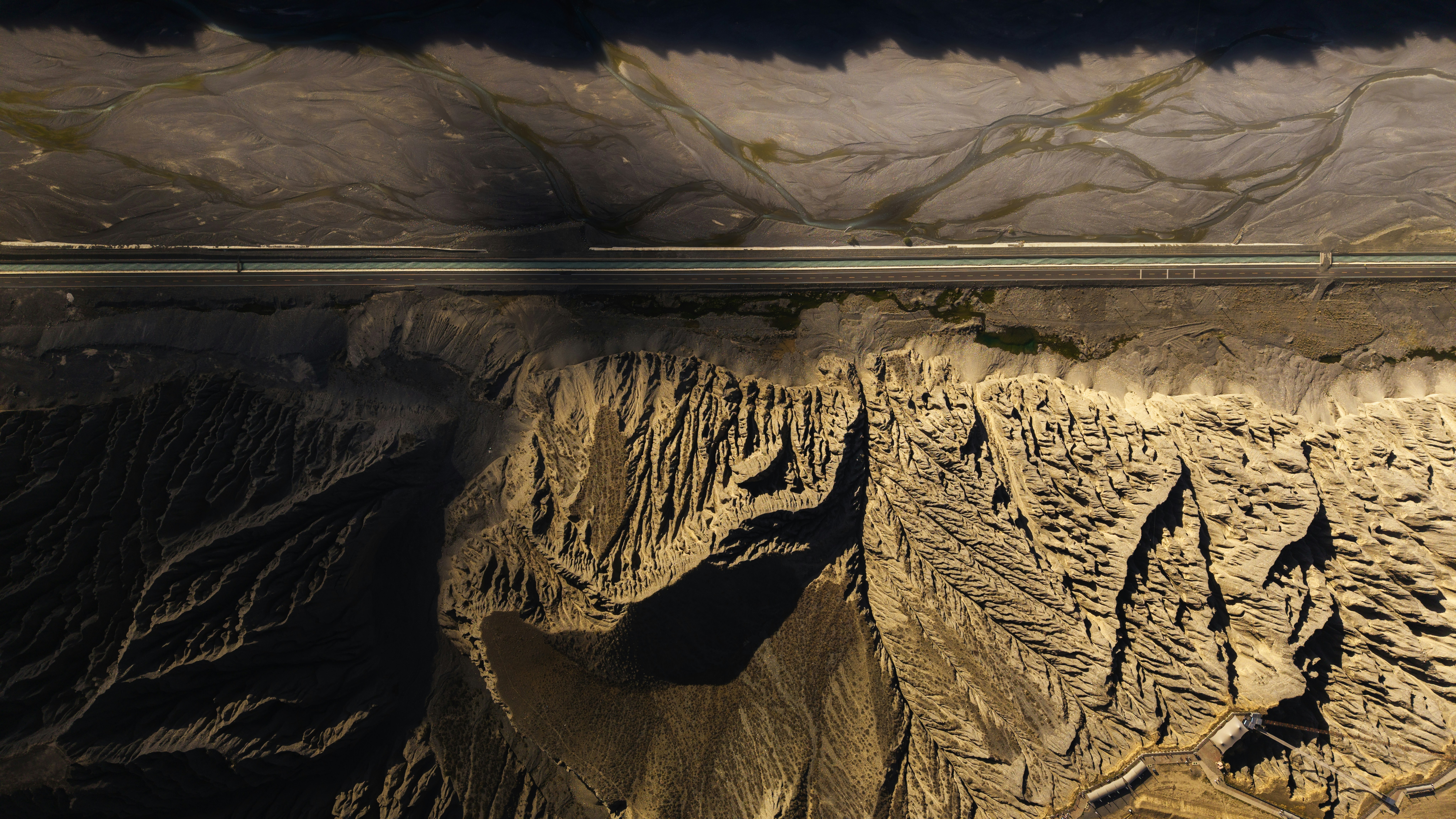 A sunlit canyon wall with intricate rock textures runs beneath a distant barrier, captured with a narrow strip of sky and a road-like line across the top. Aerial perspective highlights the erosion patterns and depth.