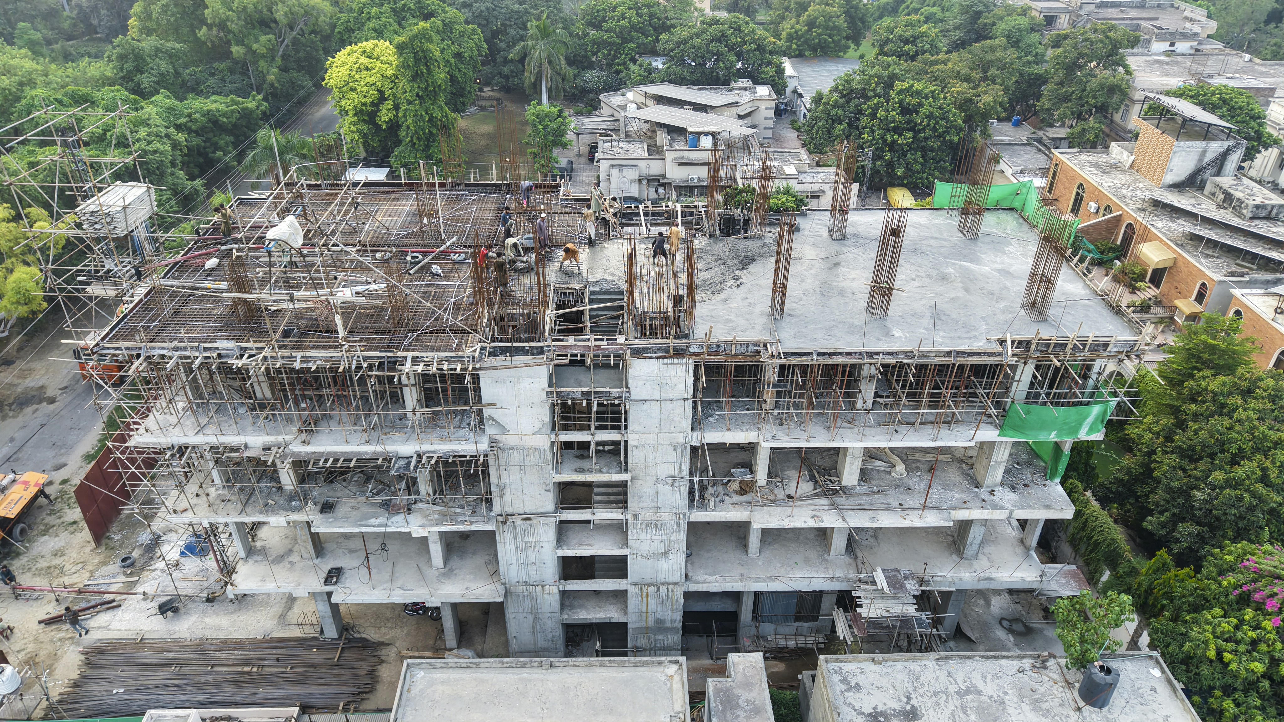 Multi-story concrete under-construction building framed with steel rebar visible amid an urban tree-lined setting. Construction site activity on a cloudy day with surrounding greenery and neighboring structures.