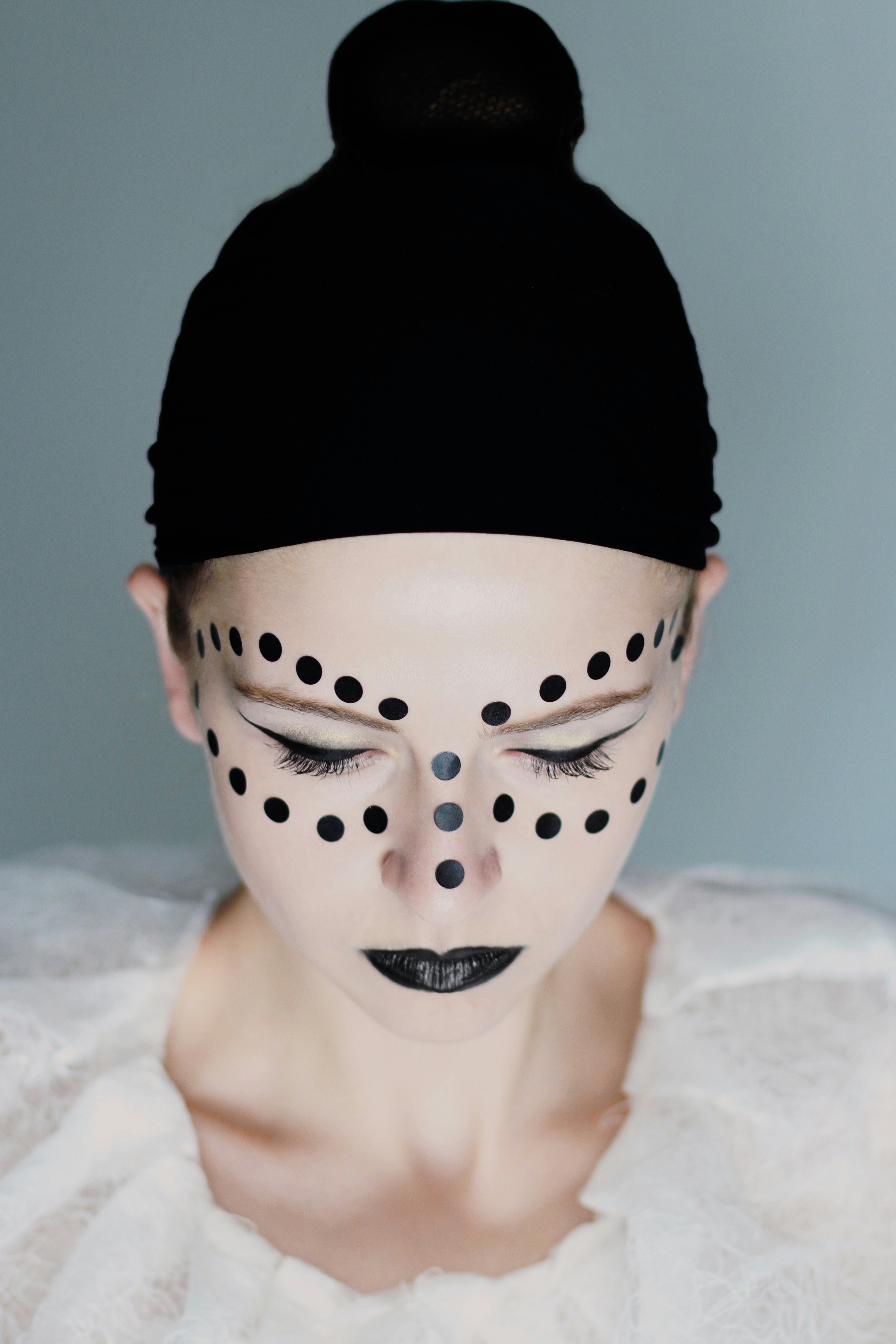 Close-up portrait of a person with bold black dots arranged in symmetrical patterns on the face, wearing a black headwrap and white textured garment, set against a muted backdrop. This is a stylized makeup or body art scene.