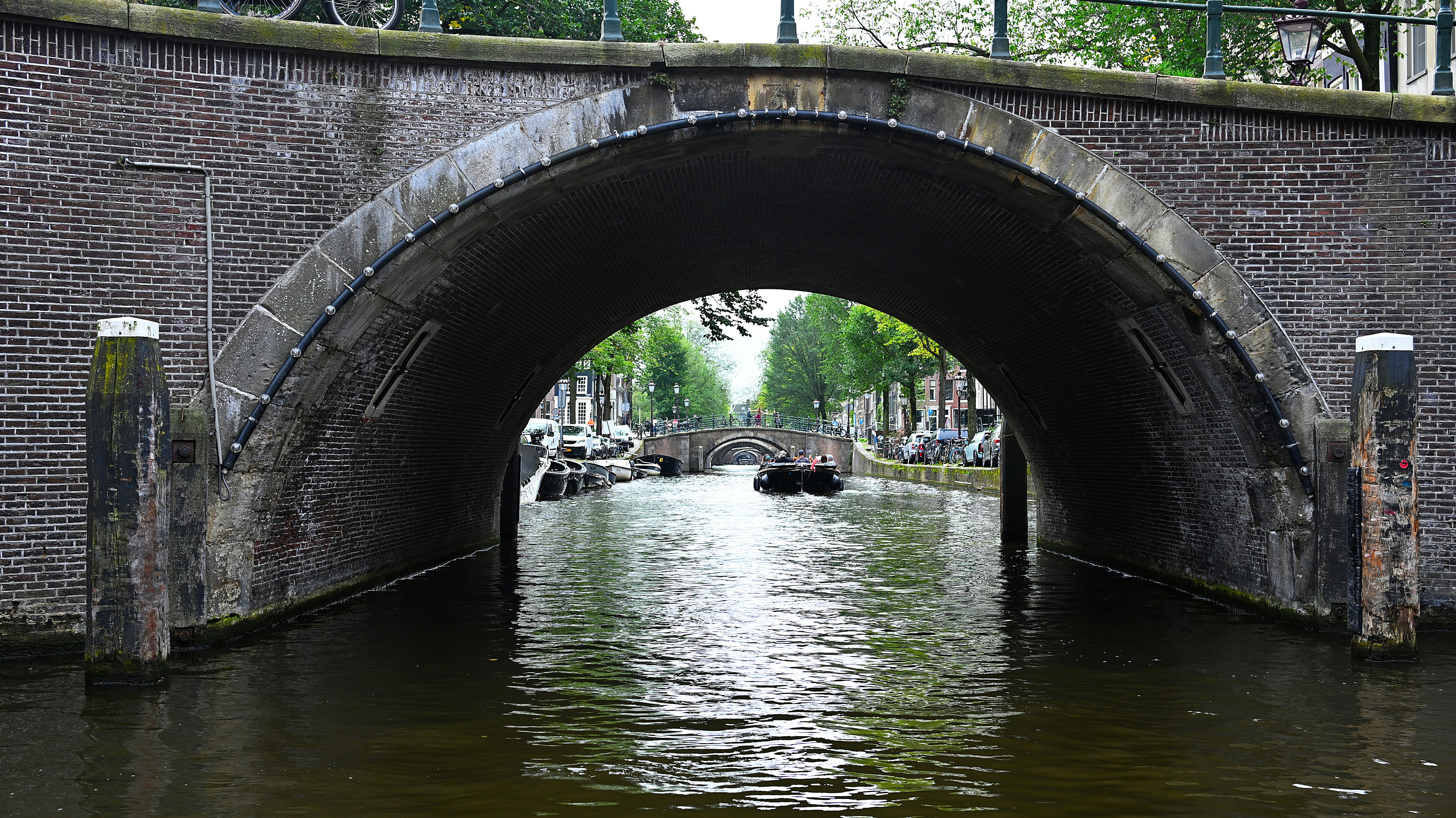 A brick arch bridge spans a calm canal with boats and trees lining the banks, creating a moody urban waterway scene.