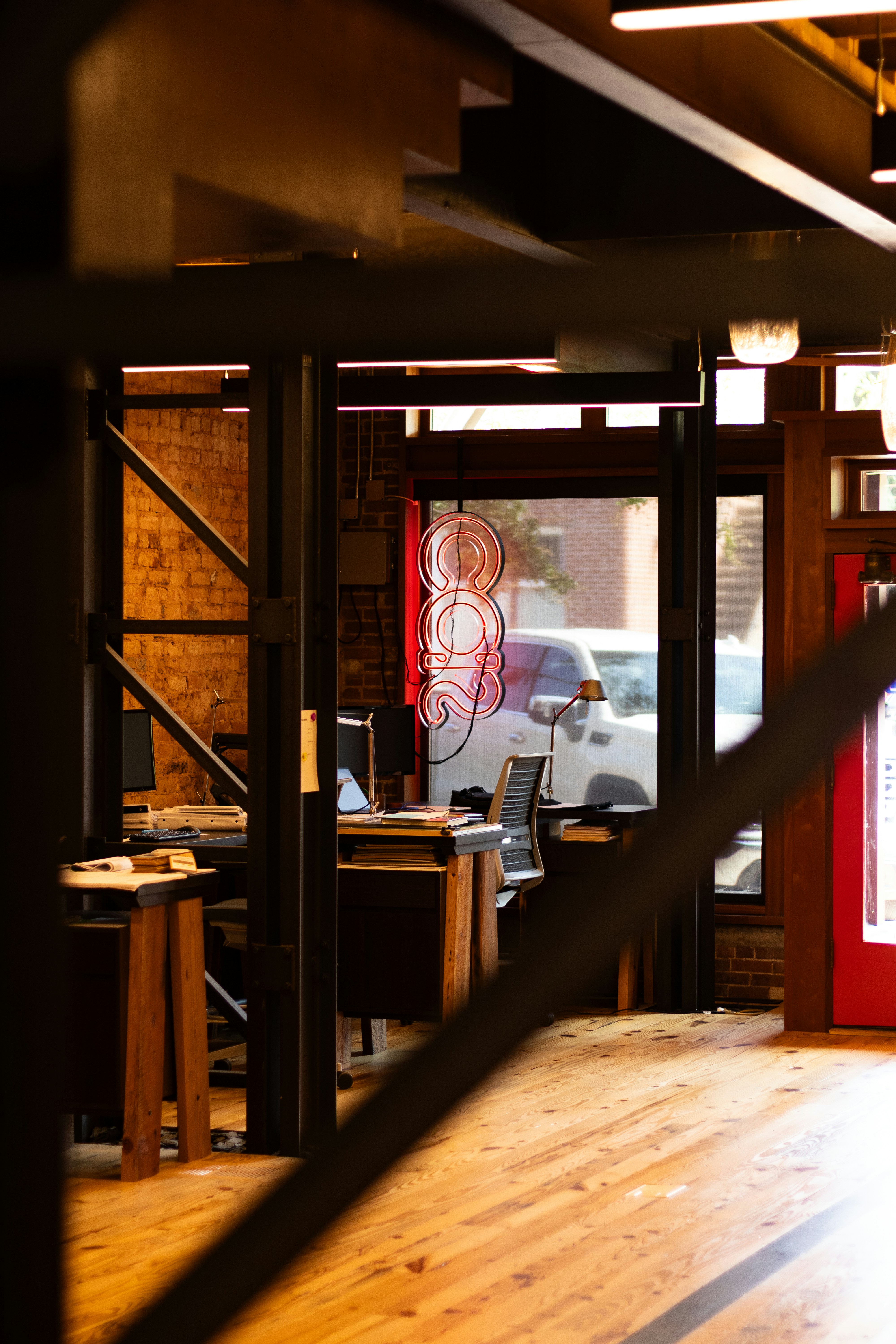 Warm interior of a cafe with wooden floors and desks, a neon sign visible through large windows near the entrance.