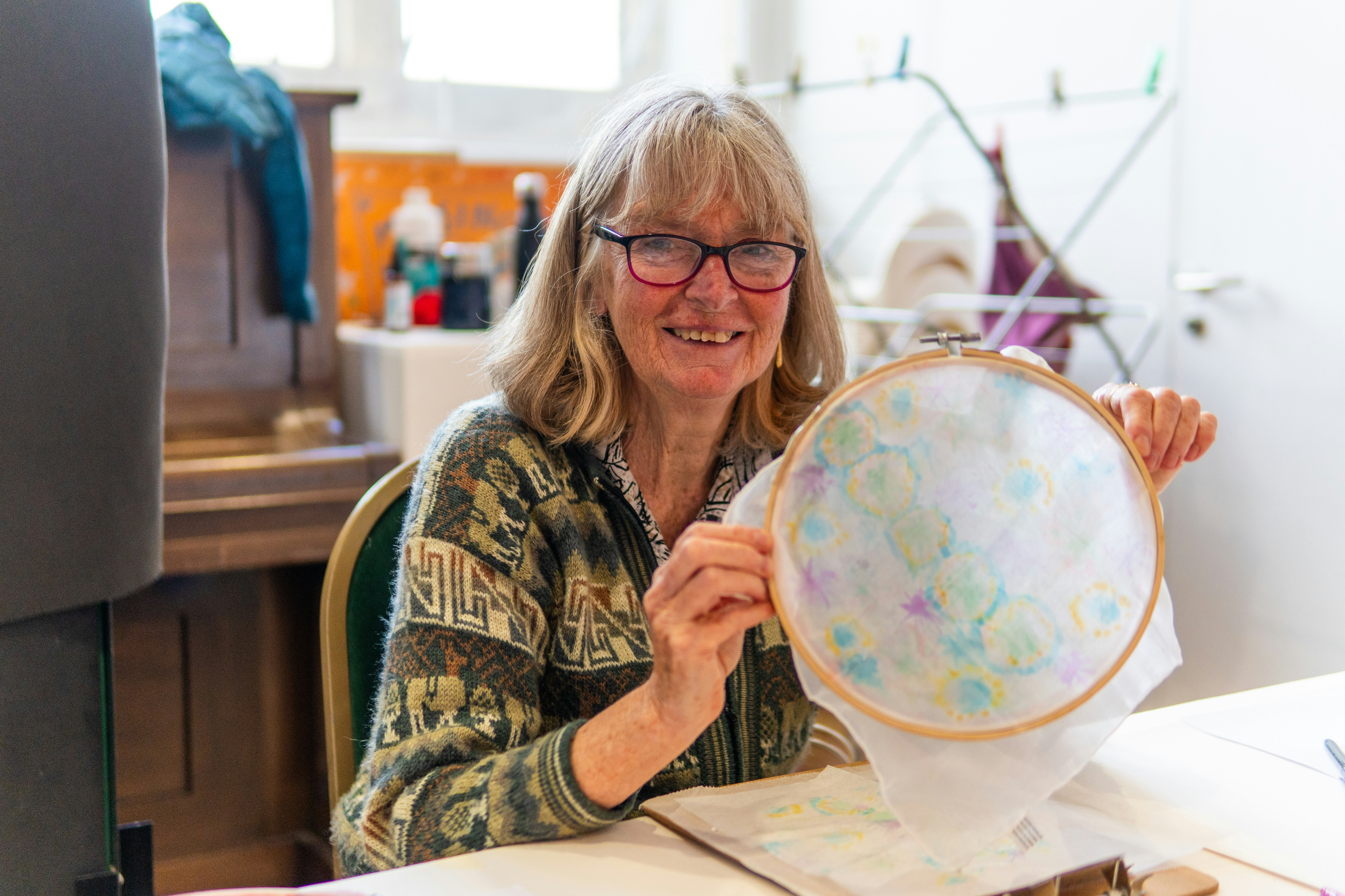 A smiling woman holds up a dyed embroidery hoop with a floral design in a cozy indoor setting.