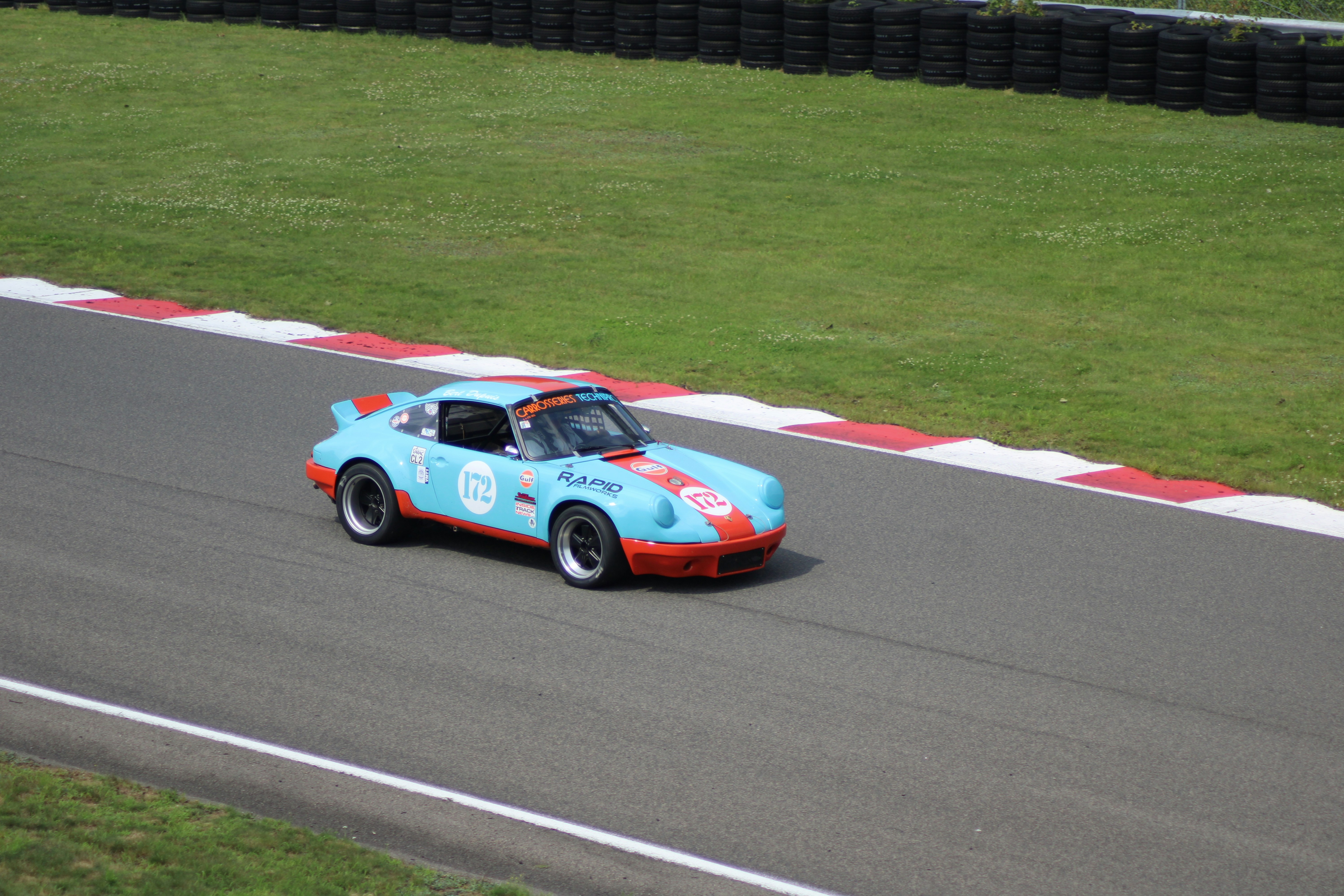 Blue race car with number 172 on a curving track, green grass and tire barriers in the background, captured mid‑motion during a race. Logo and sponsor decals visible on the body.
