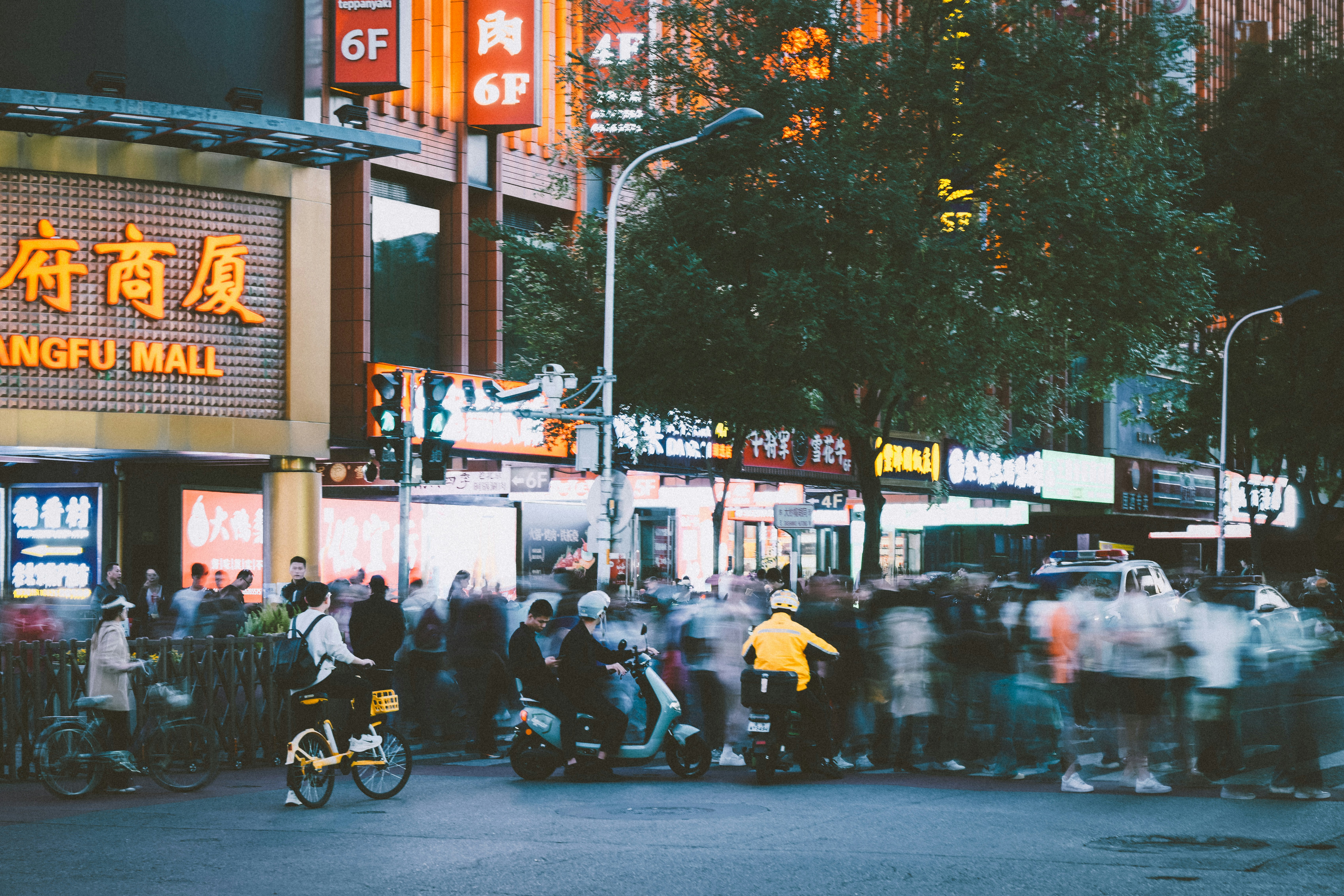 Busy urban intersection with blurred pedestrians and motor scooters under neon signage at dusk; storefronts glow, conveying a sense of nightlife energy.